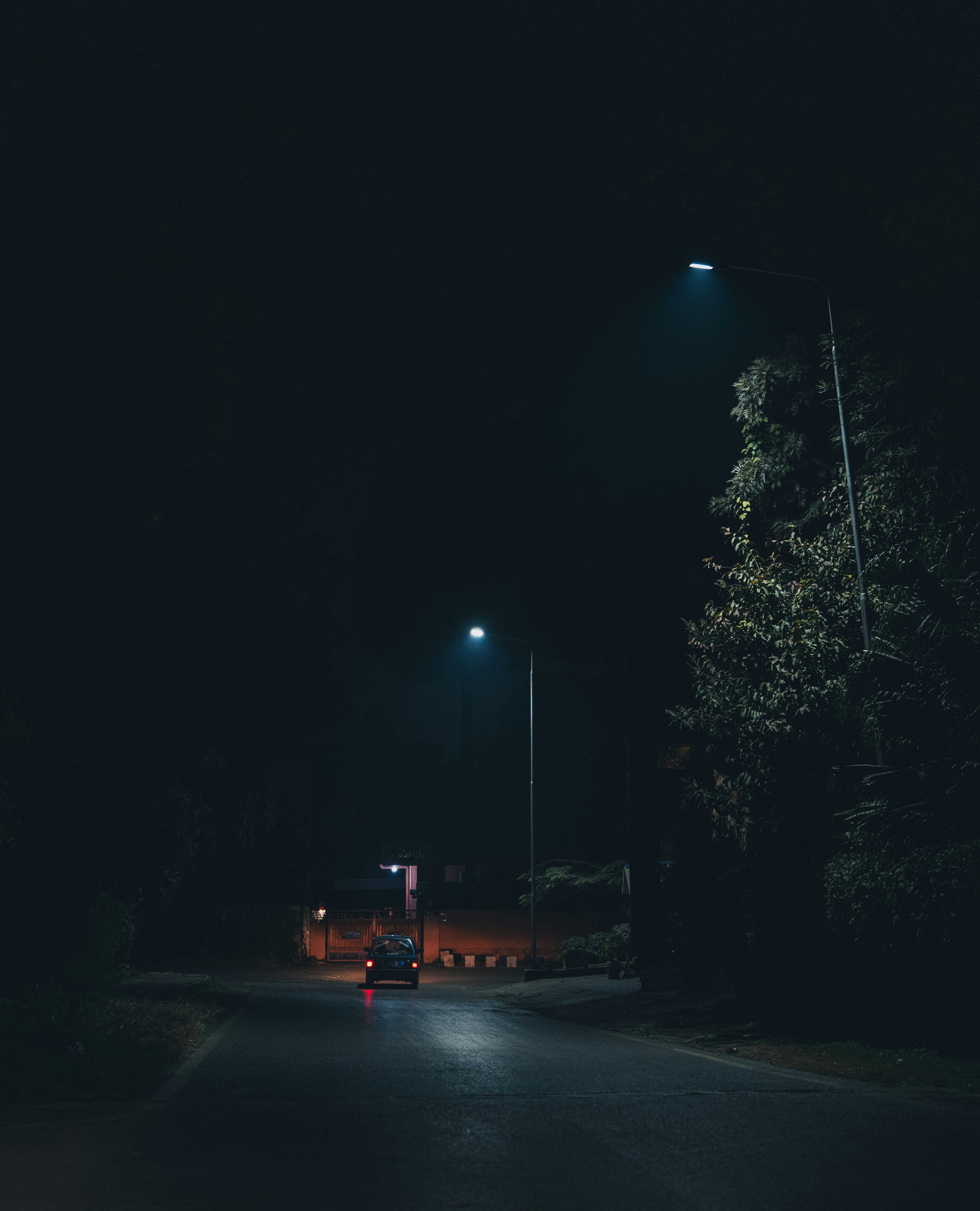 A cinematic night photograph capturing a lone car passing under glowing streetlights on an empty road. The moody blue tones and subtle orange highlight from the car create a calm yet mysterious atmosphere. Shot in Islamabad, Pakistan.