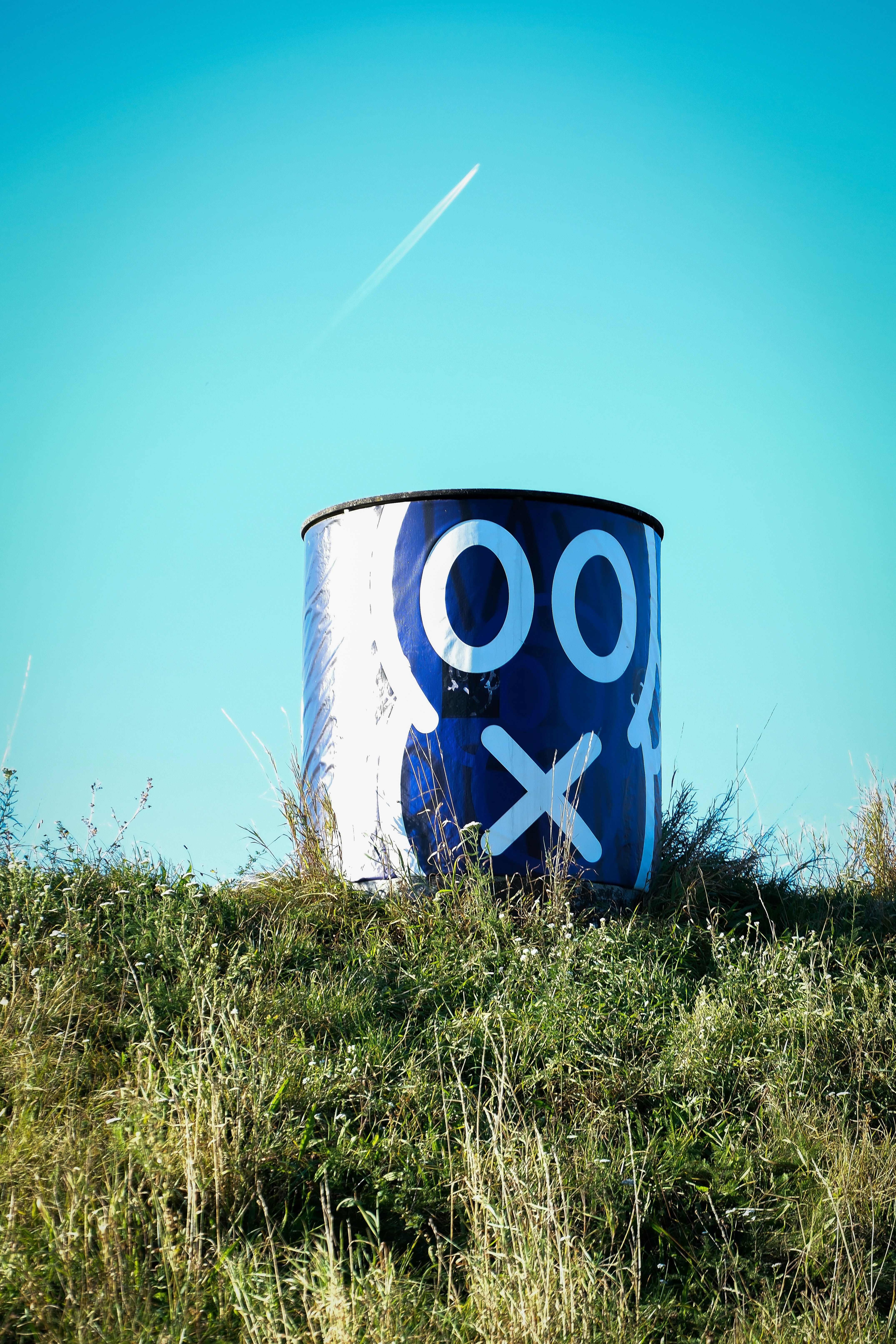 A bold blue-painted cylinder with white abstract symbols stands on a grassy hill under a turquoise sky.