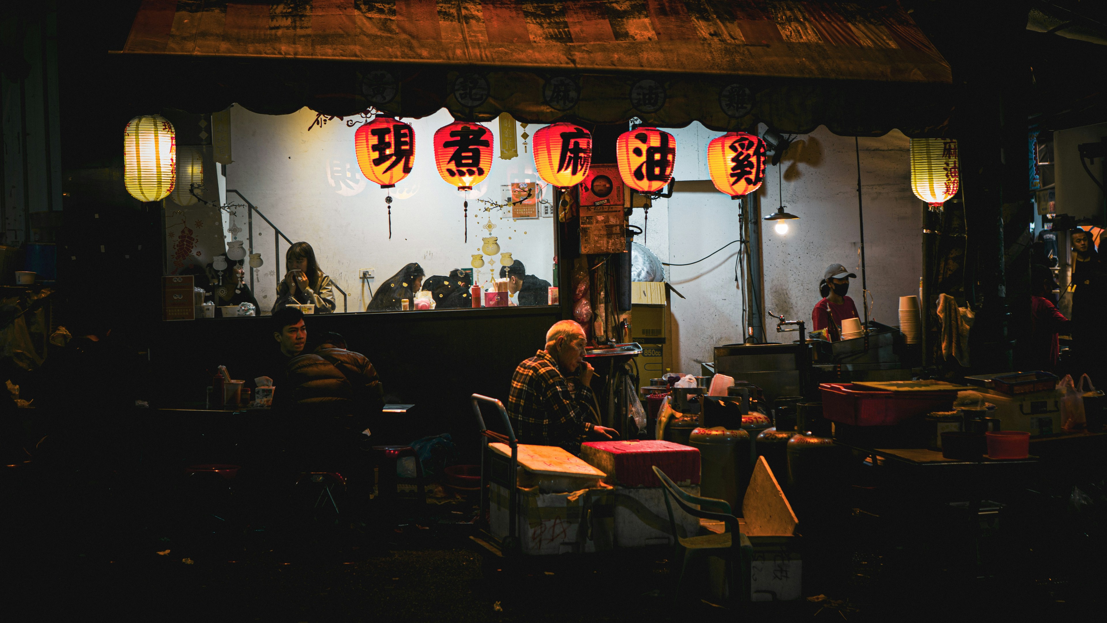 Five glowing red lanterns, advertising "freshly cooked sesame oil chicken", cast a warm, cinematic light on a humble food stall in Taichung's Feng Chia Night Market. Patrons, including an older man in a plaid shirt, gather at simple tables, immersed in the intimate, communal atmosphere.