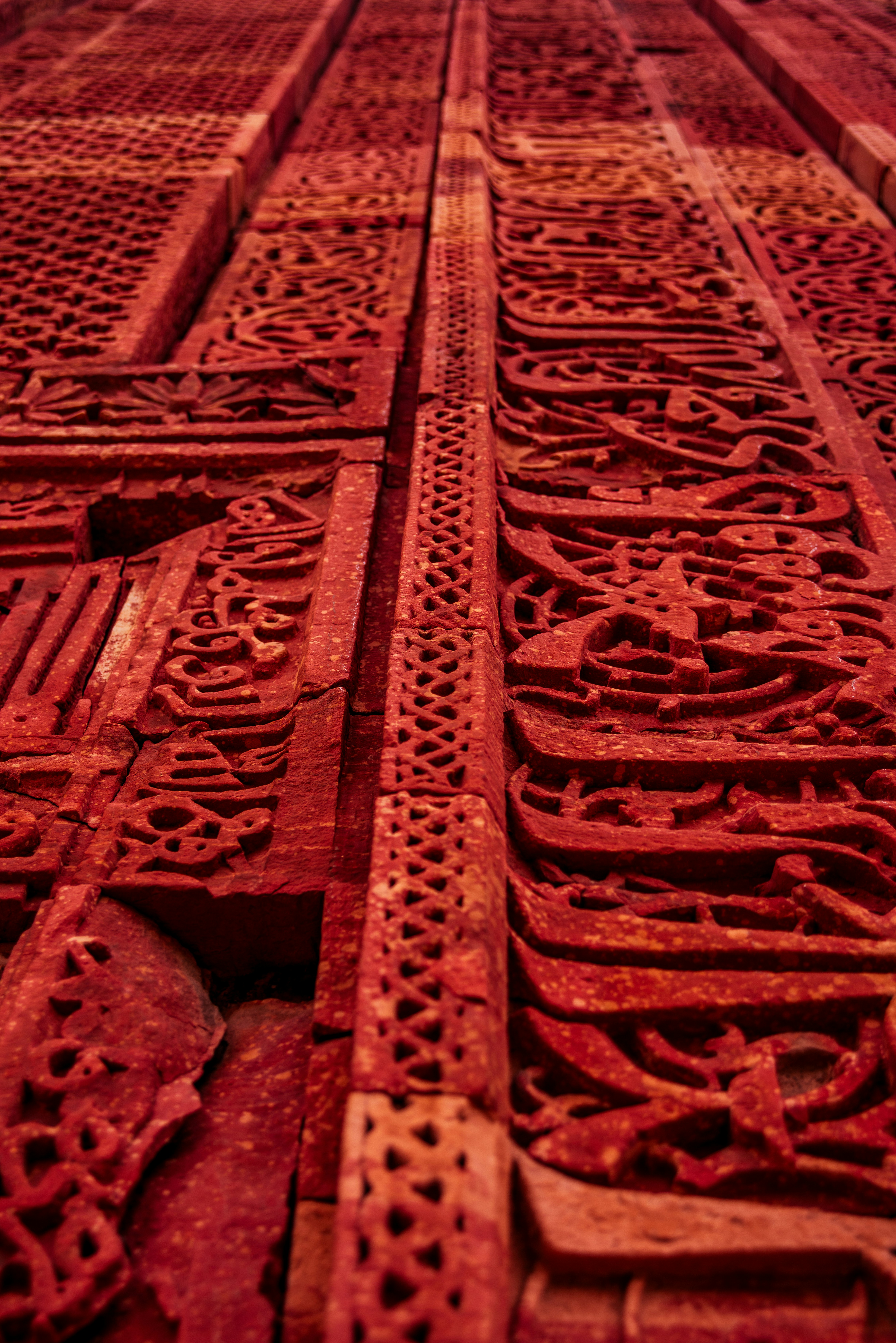 This extreme close-up of deeply carved red sandstone reveals intricate Islamic calligraphy and geometric patterns, emphasizing the weathered texture and rich, warm tones. The upward perspective creates a powerful sense of height and verticality. The texture is perfect for use as an architectural background, an overlay to add historic depth to a design, or as a website element to evoke a sense of heritage and craftsmanship.