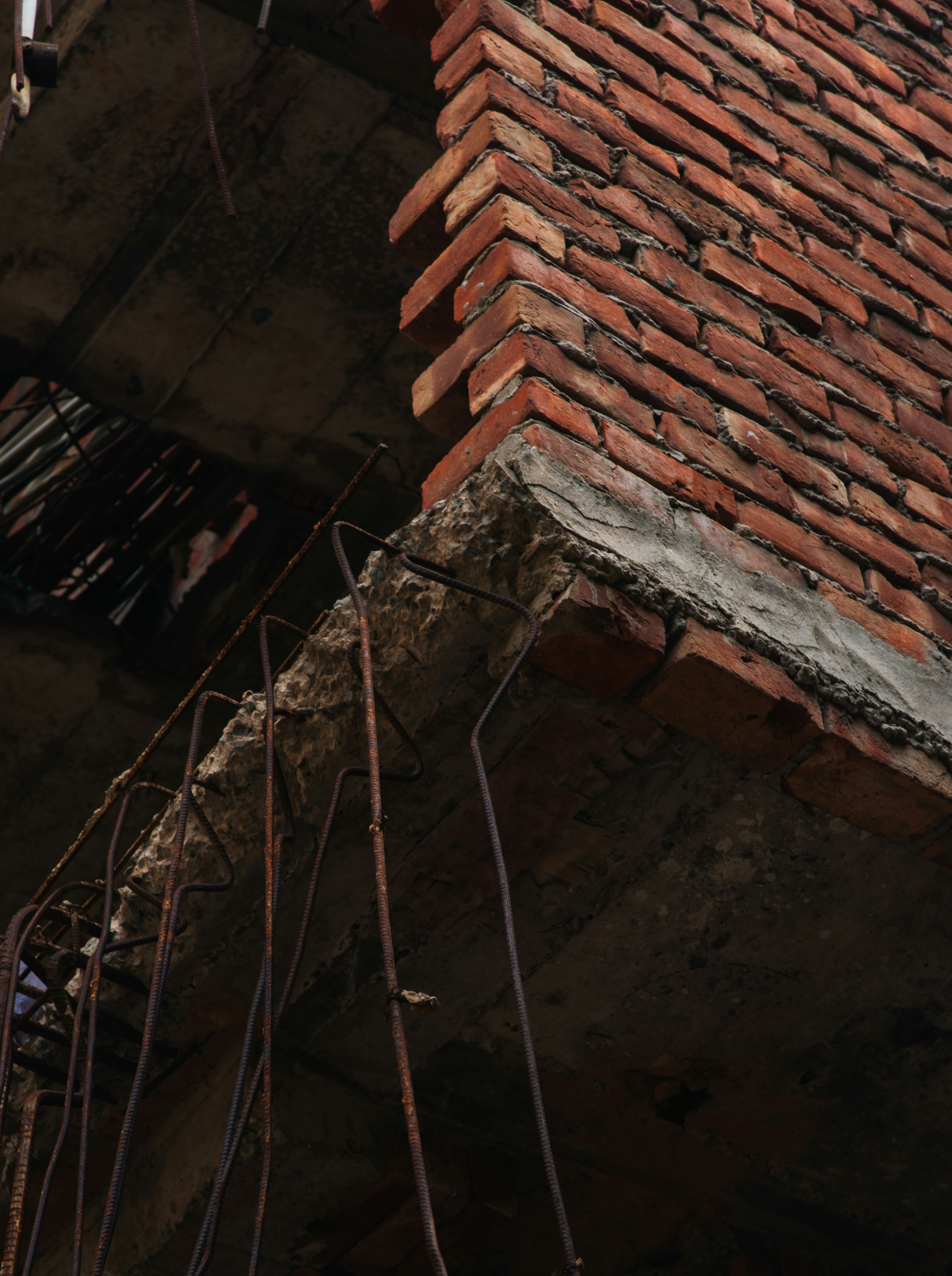 This image captures the raw, gritty texture of urban decay and ongoing construction, dominated by an angled corner of a wall with weathered red bricks. The foreground features exposed, rusted rebar and rough, chipped concrete, emphasizing contrast and imperfection.
