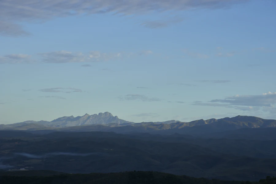 Layered mountain ranges stretch across the horizon beneath a clear blue sky with thin clouds. Soft evening light catches the distant peaks while the foreground remains in shadow.