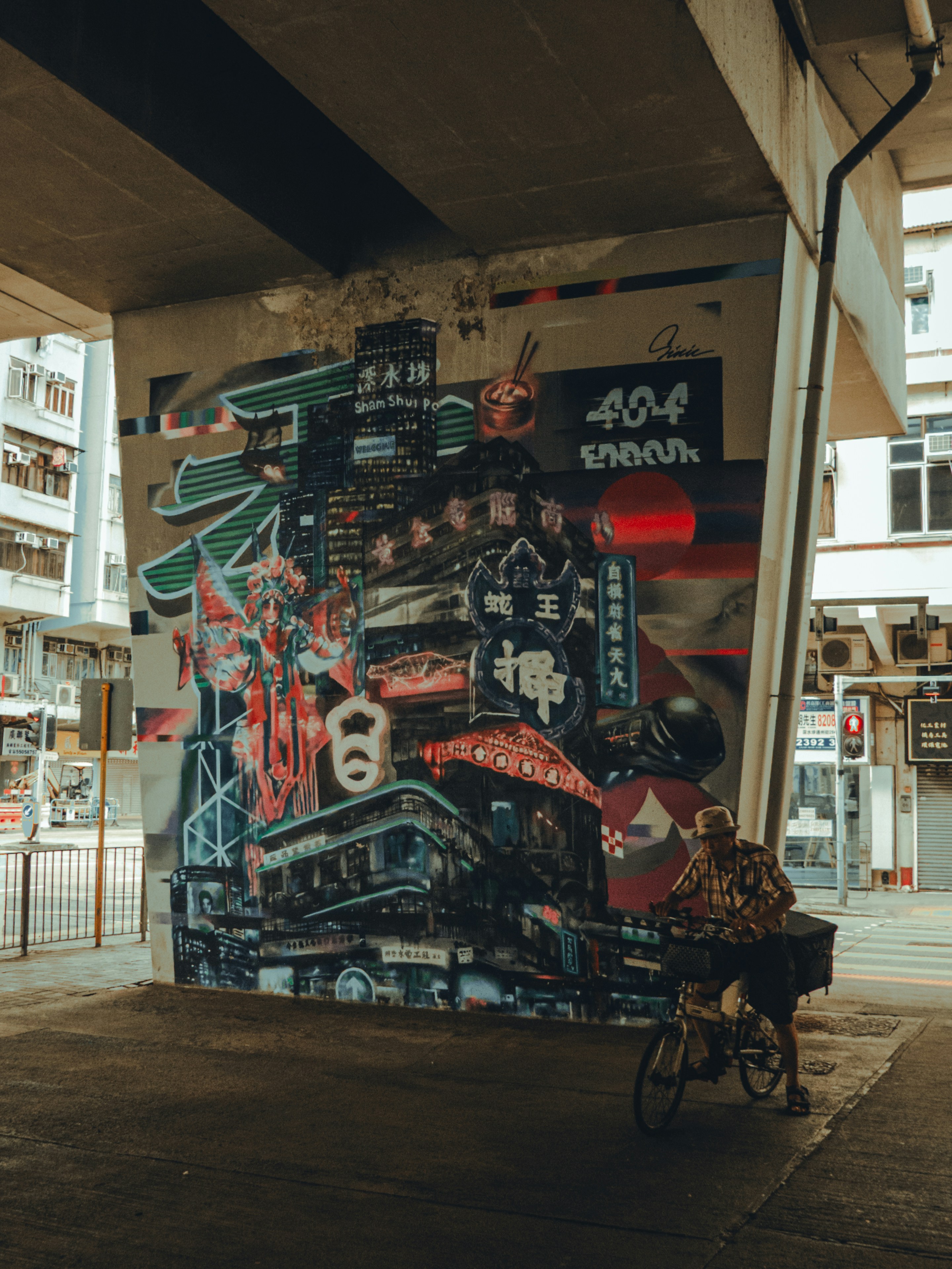 A man pushes his bicycle under a highway overpass in Hong Kong's Sham Shui Po, dwarfed by a massive, vibrant graffiti mural. The artwork, a chaotic collage of neon signs, local buildings, and cyberpunk "404 ERROR" text, celebrates the district's gritty energy.