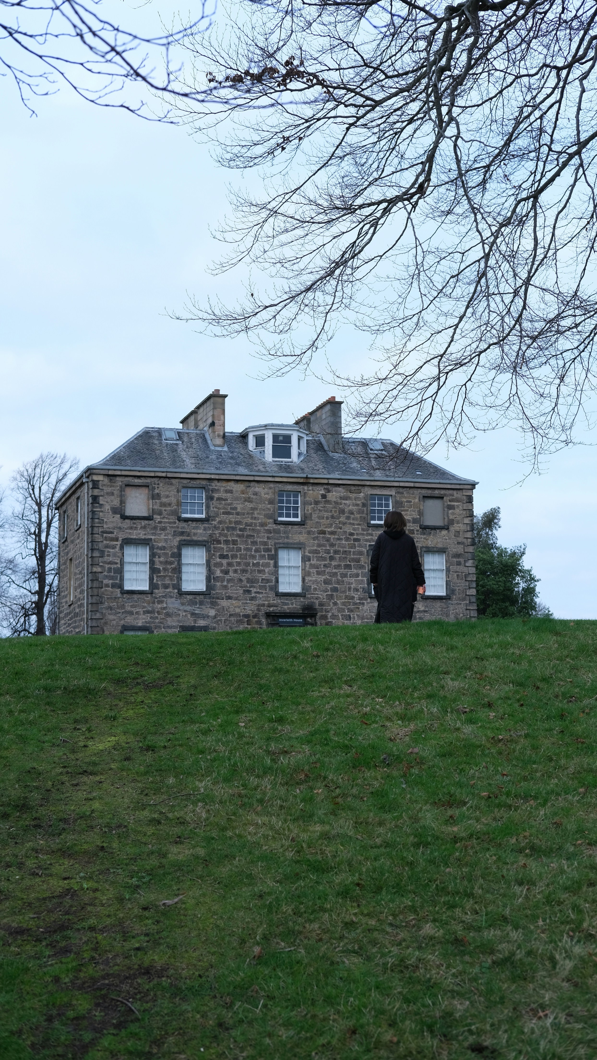 Silhouette on the Hilltop: A lone figure beside a stone manor under bare branches