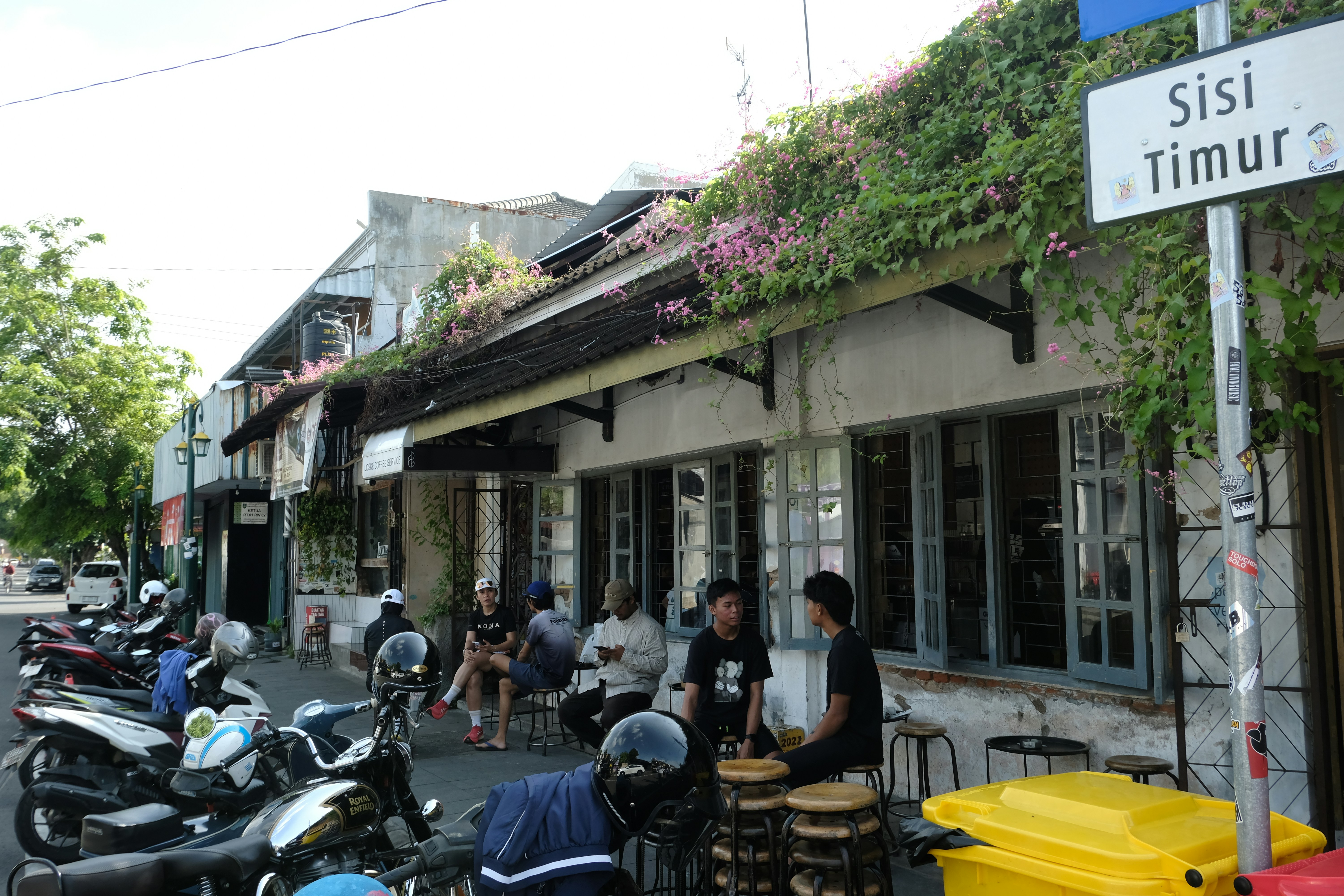 A cozy street-side café in Surakarta, Indonesia, where people enjoy casual conversations under a vine-covered roof. The sunlight, rustic building, and rows of parked motorbikes create a relaxed local atmosphere typical of Indonesian coffee culture.