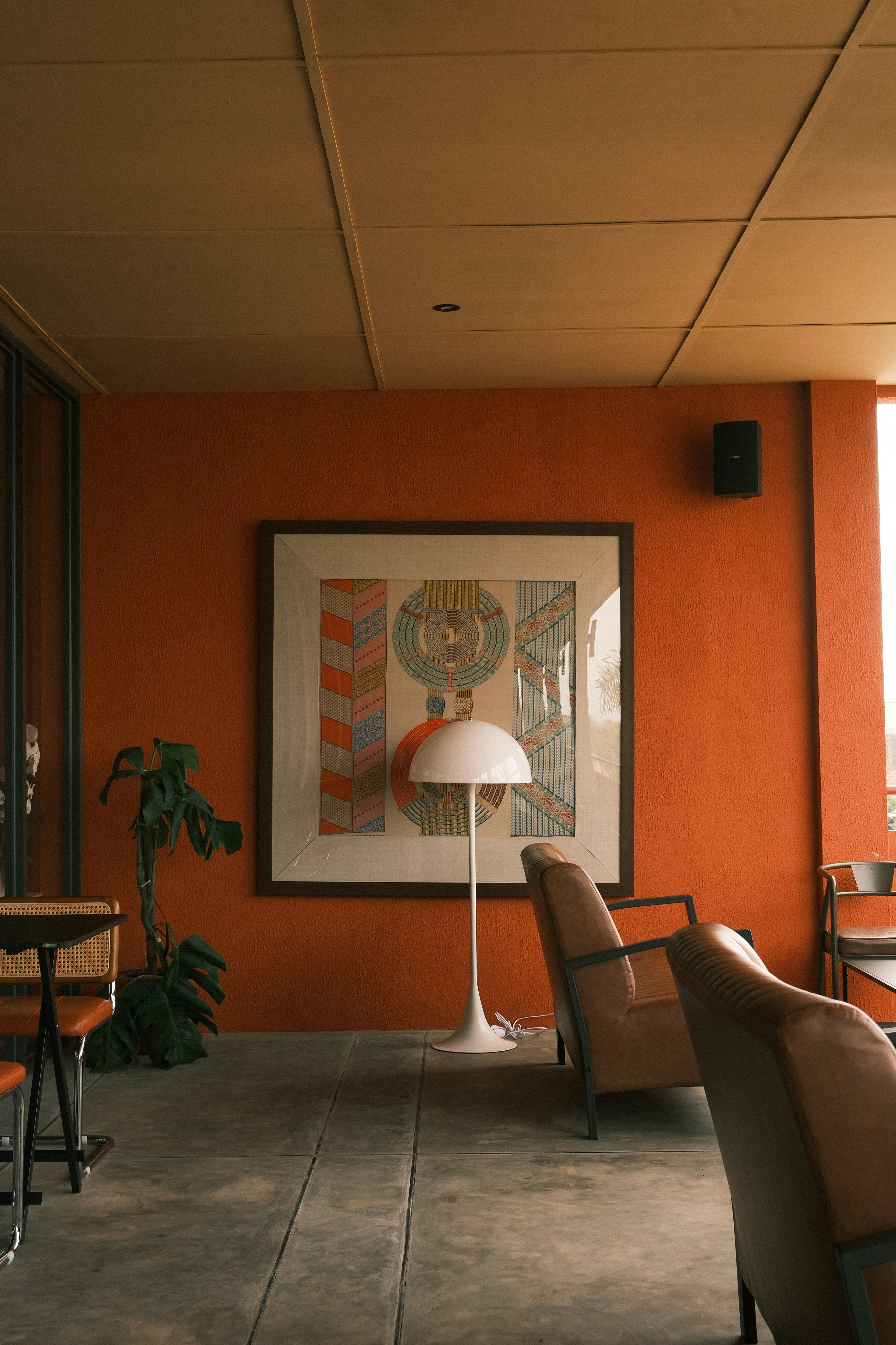 A cozy corner featuring a stylish floor lamp, a vibrant geometric artwork, and contemporary furniture, set against a bold orange wall in Malang, Indonesia.