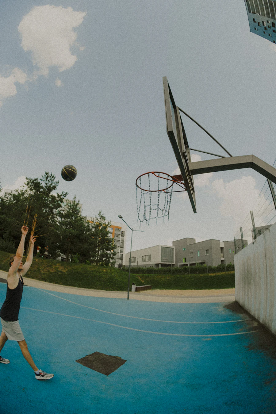 Player releasing a basketball toward a rim above a bright blue outdoor court with residential buildings and trees in the background.