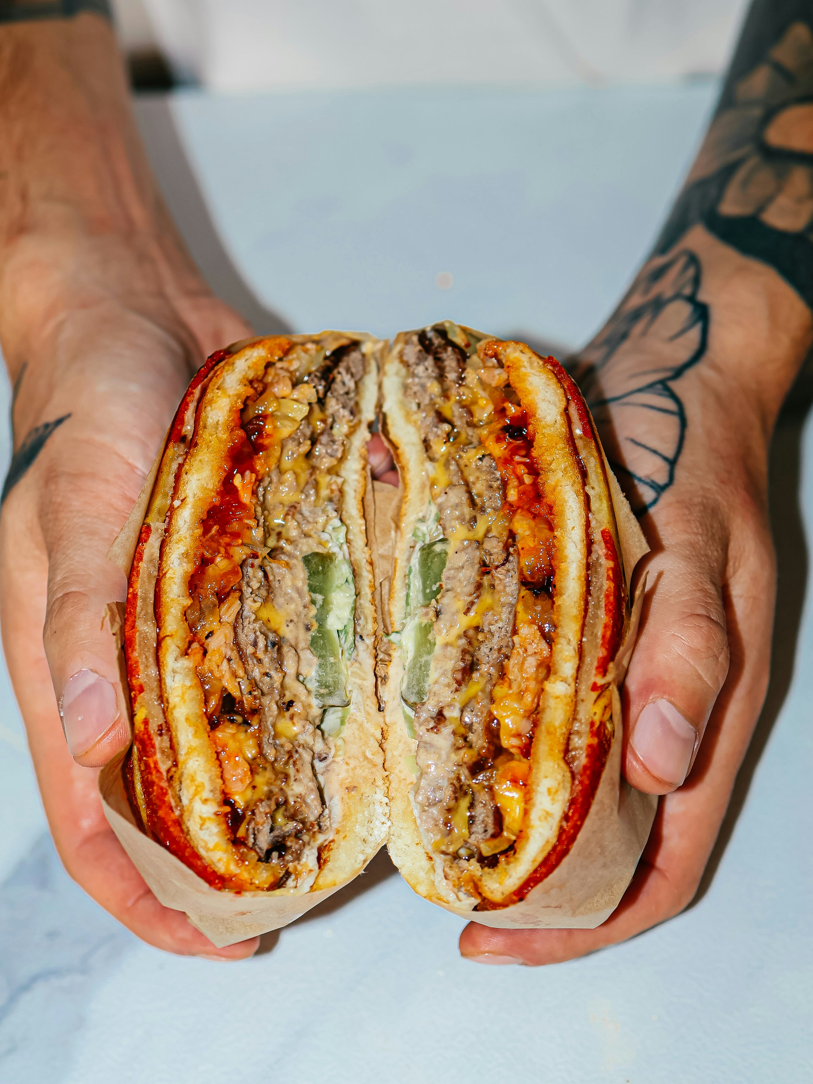 Ink and Layers: Two Tattooed Hands Cradle a Hearty Layered Sandwich