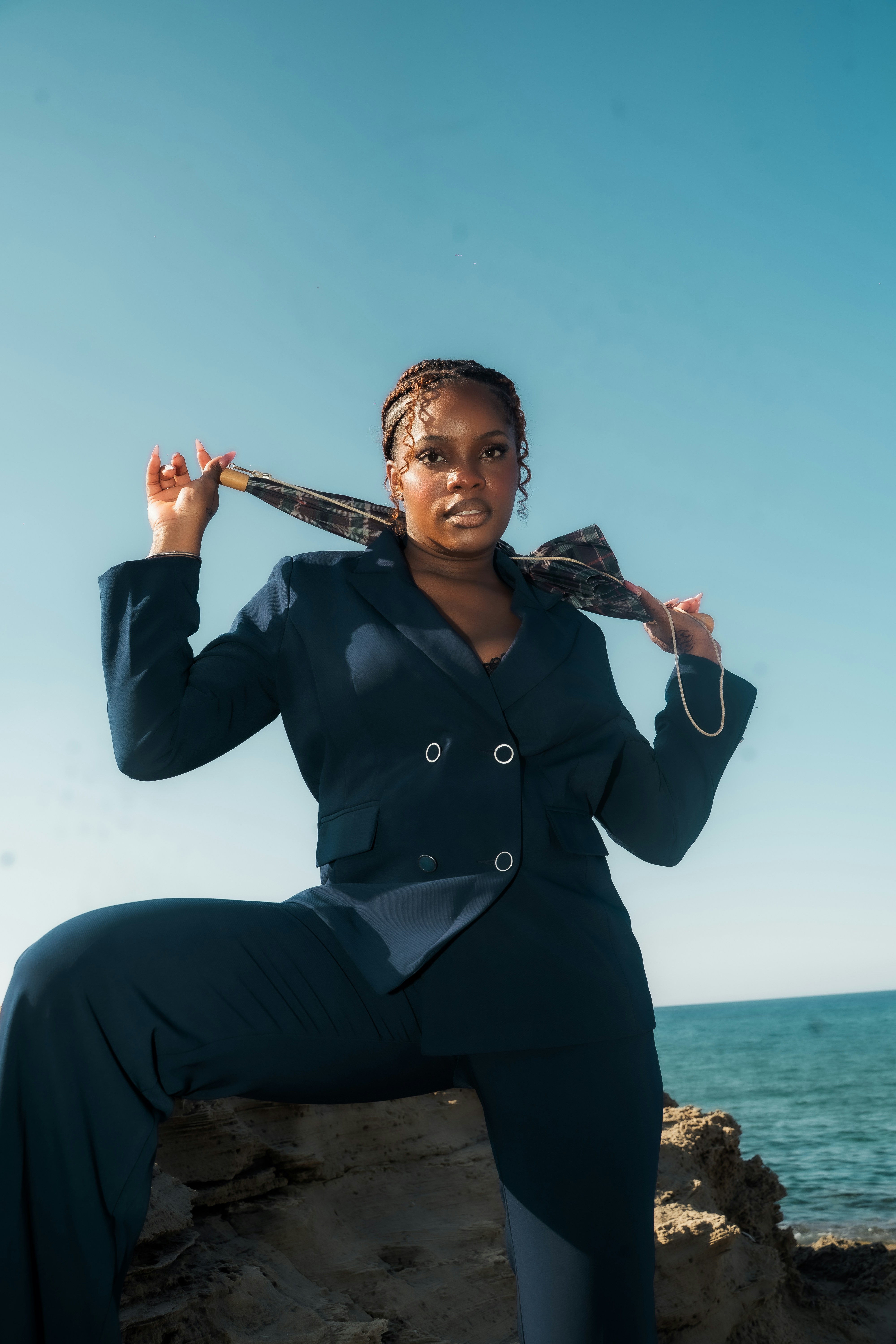 A vibrant, empowering portrait of a woman in a navy blue suit posing with attitude near the beach. The photo captures confidence, freedom, and individuality against a bright coastal backdrop — a fusion of style and energy under clear blue skie