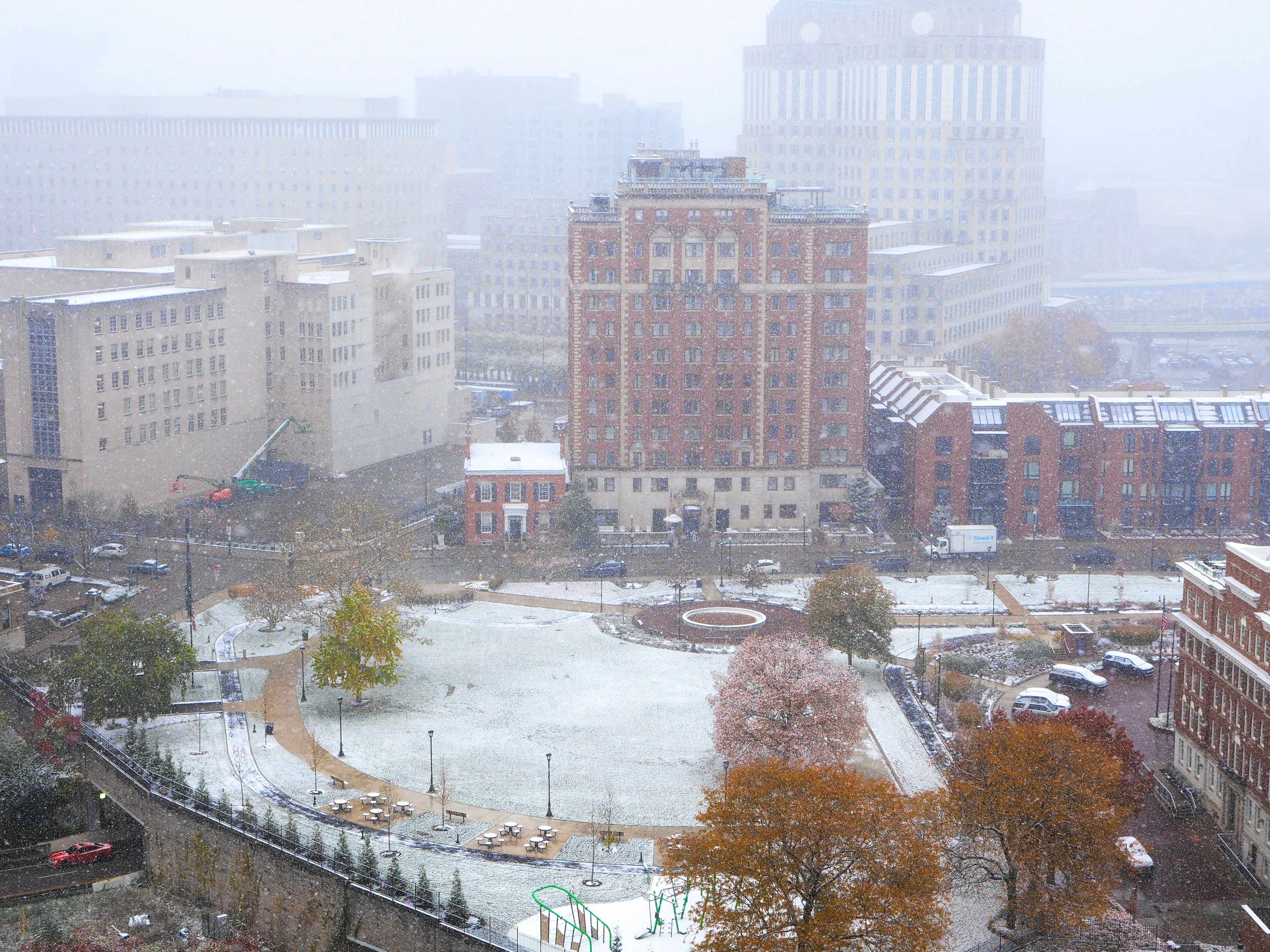 The first snowfall of the season hits downtown Cincinnati while some stubborn fall trees are still holding onto their leaves. The view looks down on Lytle Park surrounded by old brick buildings and newer high-rises, everything getting covered in white while the snowfall gets heavier. That weird in-between moment when you're not quite done with fall but winter shows up anyway.