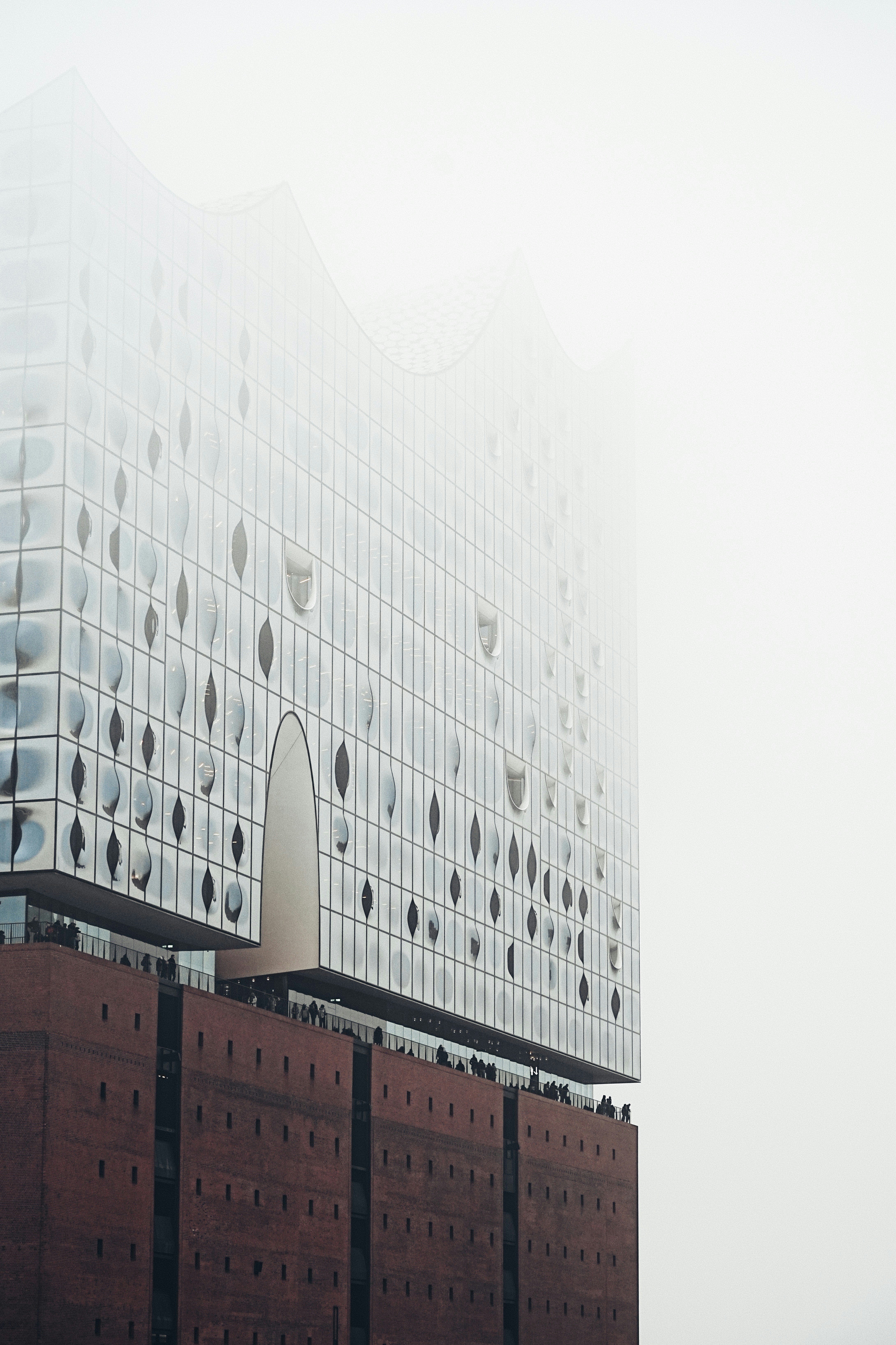 The image shows a modern architectural building with a striking glass façade, partially obscured by fog. The structure rises above a brick foundation, blending contemporary design with traditional materials. The fog creates an ethereal atmosphere around the building.