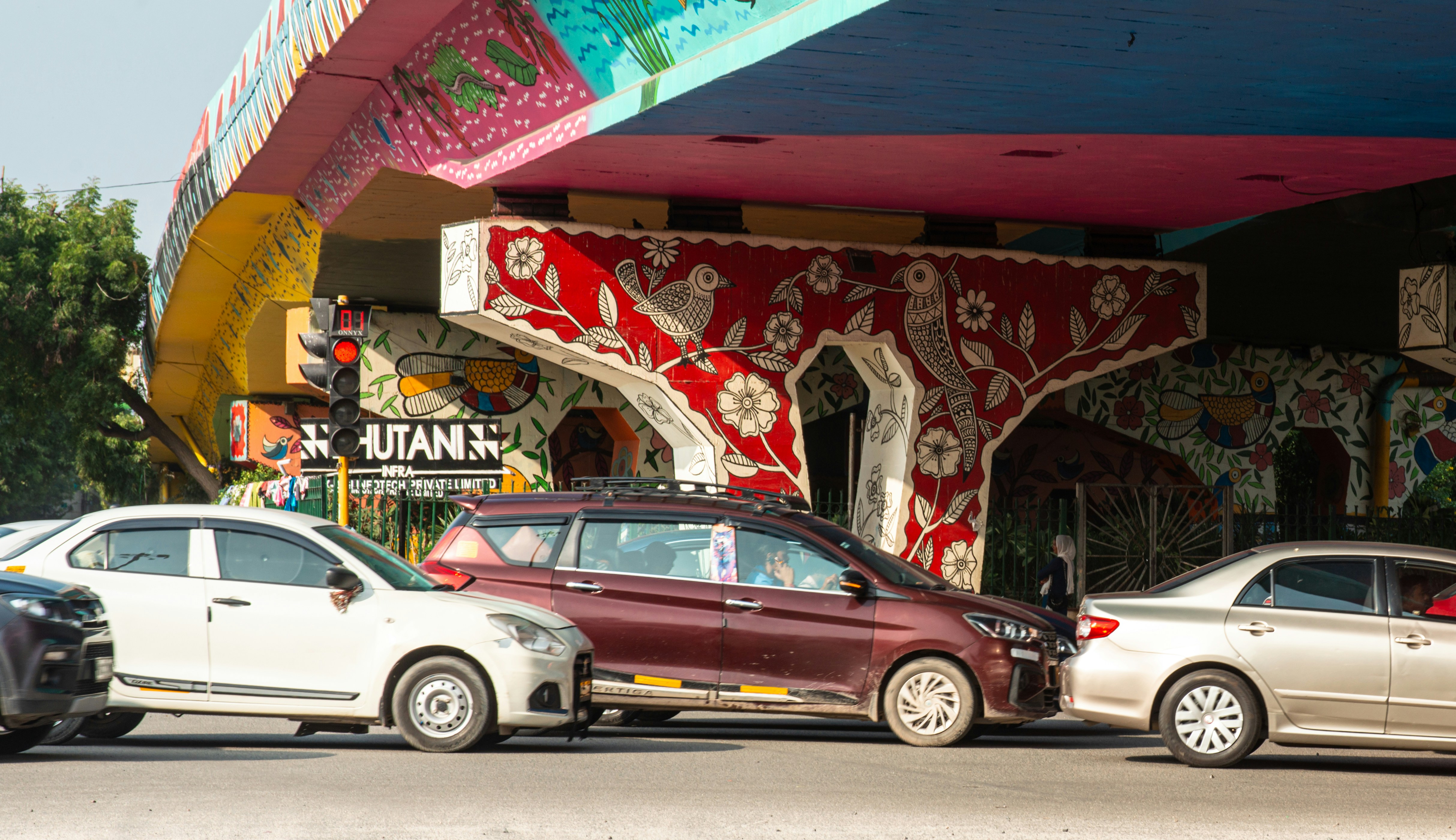 A colorful painted flyover in Delhi, India, featuring vibrant Madhubani-style art under the bridge with cars passing below. The mural showcases fish, birds, and floral motifs in red, blue, yellow, and green, reflecting India’s street art culture and efforts to beautify public spaces.