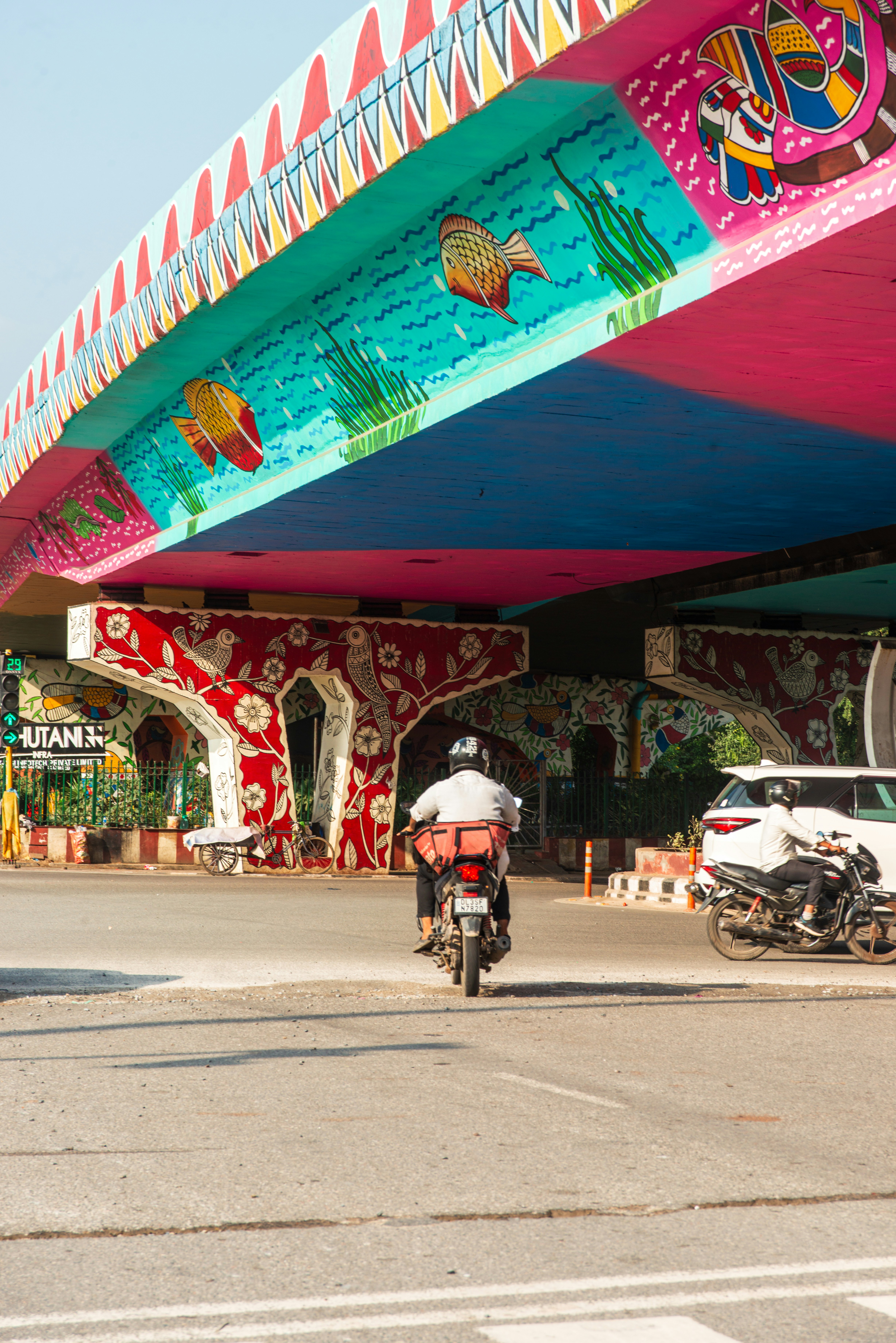 A colorful painted flyover in Delhi, India, featuring vibrant Madhubani-style art under the bridge with cars passing below. The mural showcases fish, birds, and floral motifs in red, blue, yellow, and green, reflecting India’s street art culture and efforts to beautify public spaces.
