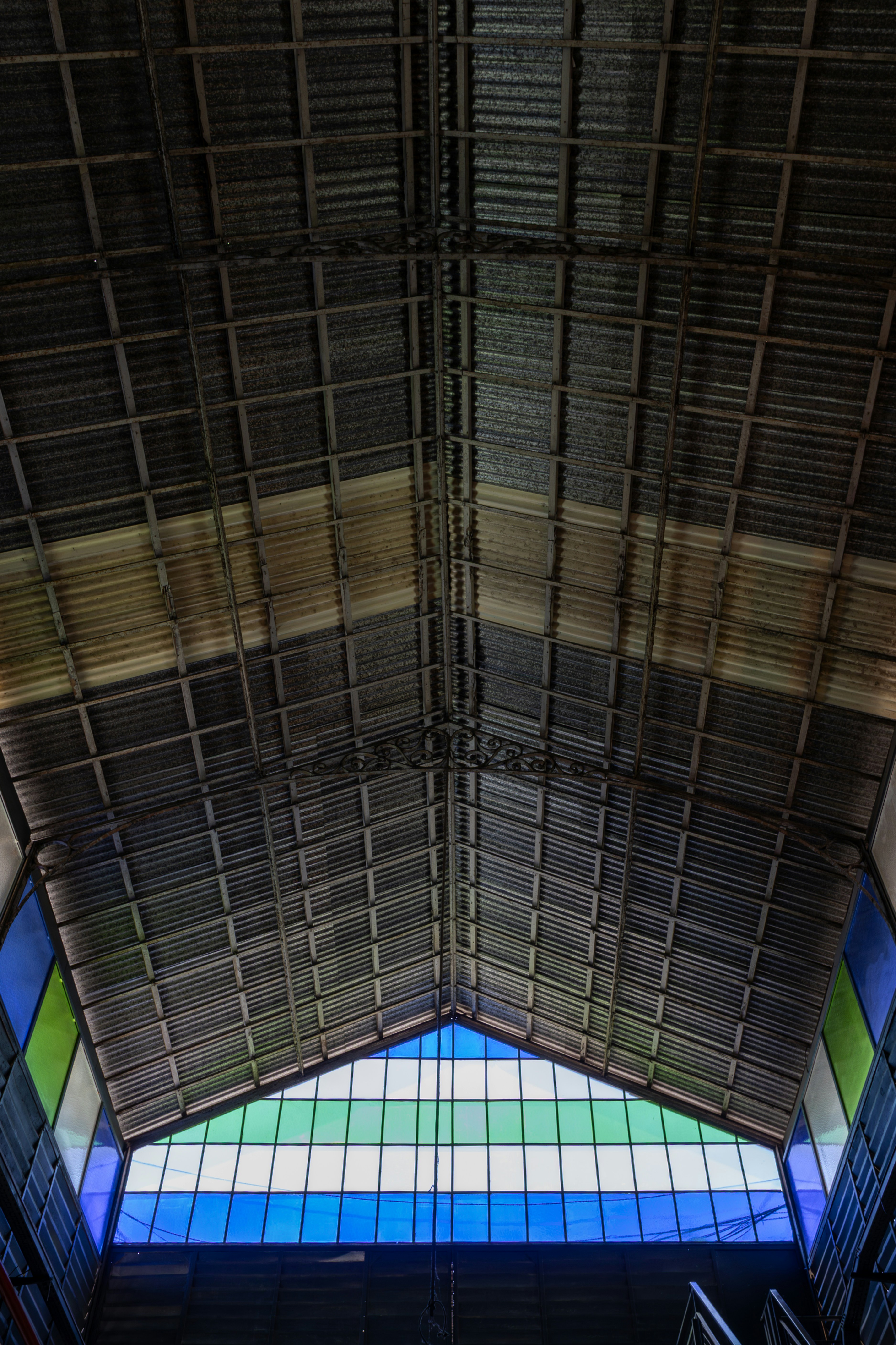 Looking up at a high, symmetrical metal grid ceiling meeting vibrant blue and green stained glass windows. An industrial, architectural perspective.