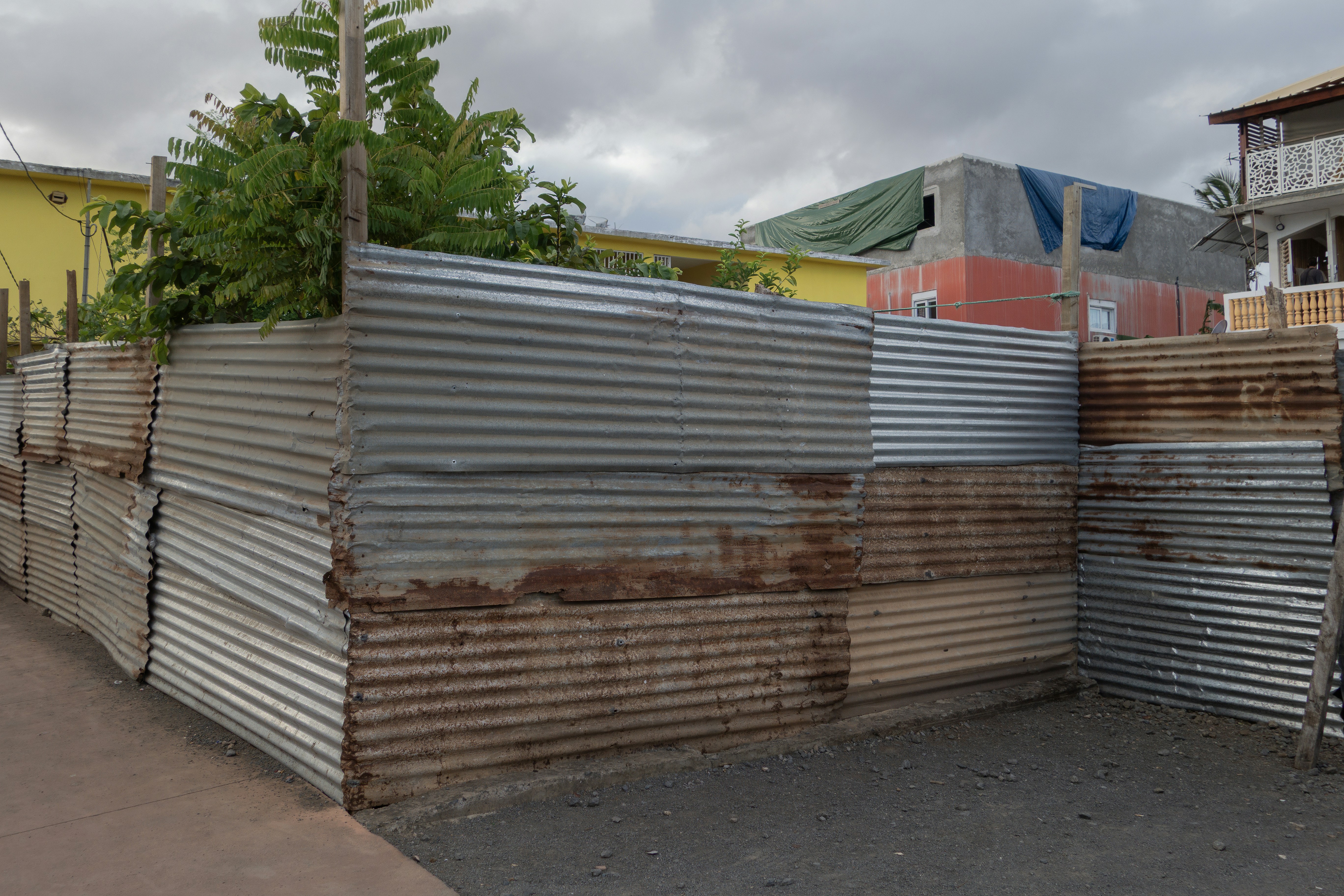 Weathered corrugated metal fences line a dusty street, with yellow houses peeking beyond beneath a gray, overcast sky.