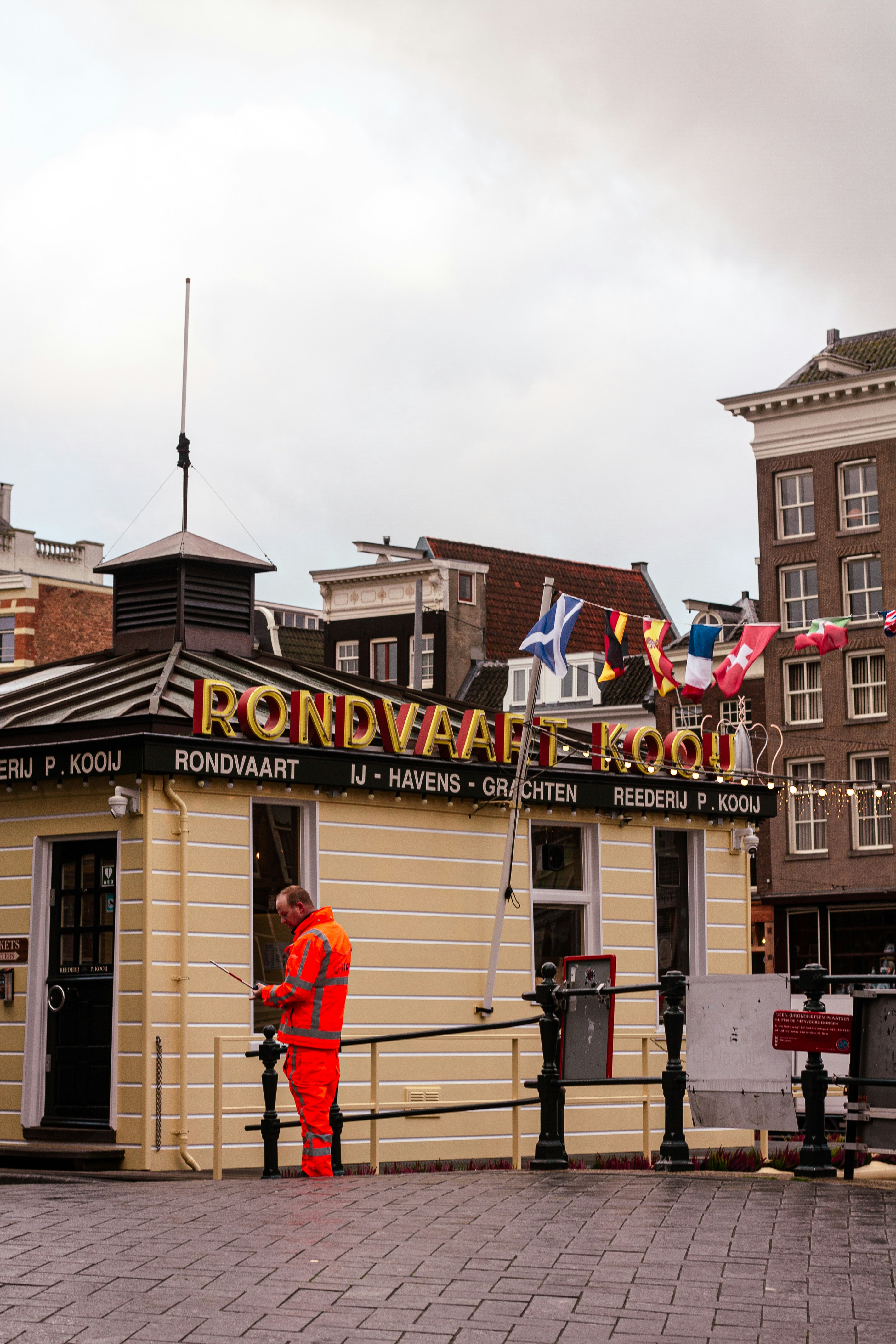 Orange Sentinel by a Canalfront Kiosk under Flag-Adorned Gables