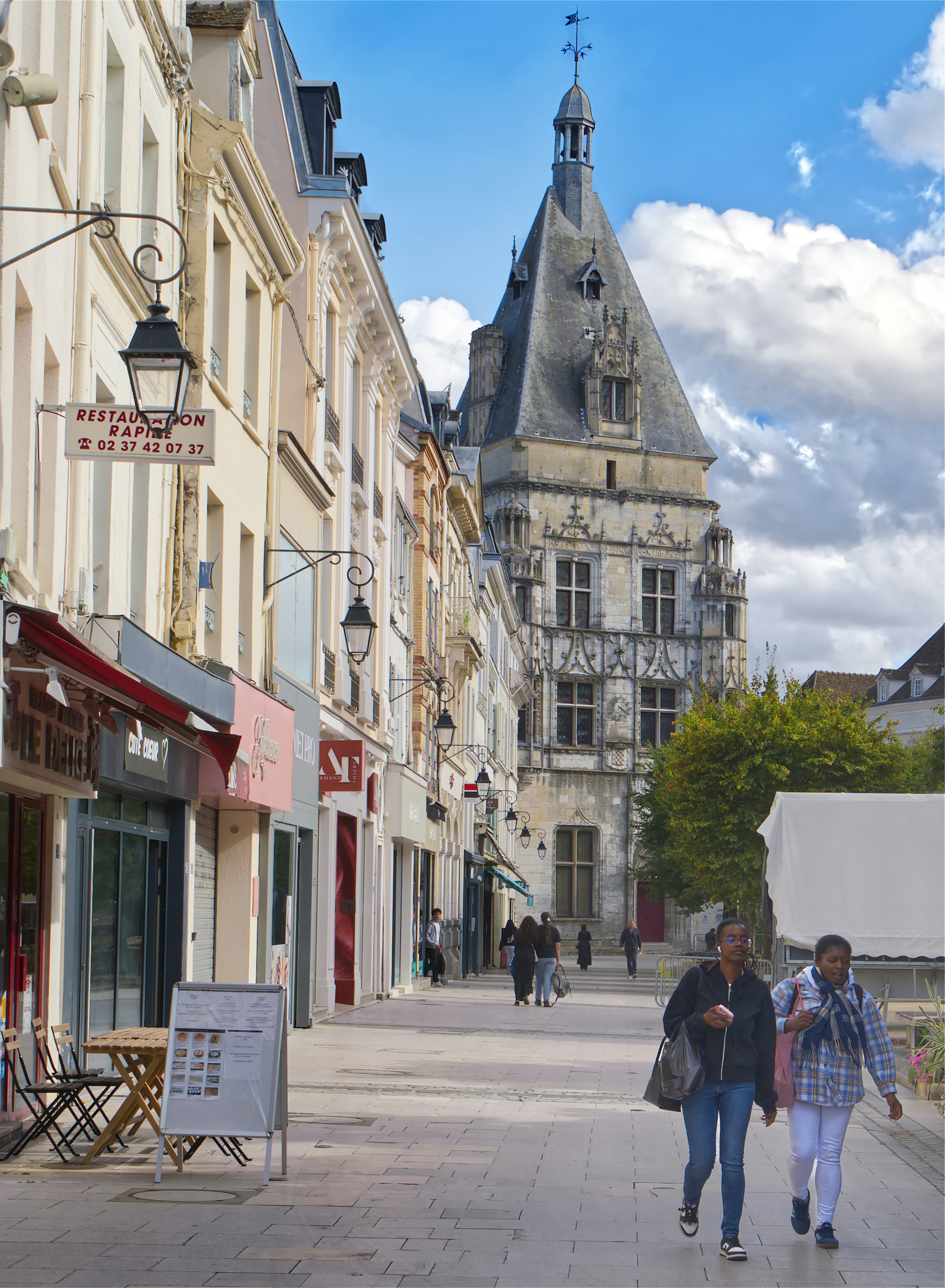 Cet après-midi, la lumière joue avec les façades de la rue piétonne, révélant chaque détail de pierre et de zinc sous un ciel à la fois doux et changeant. Au loin, l’imposant Beffroi veille sur la ville de Dreux, témoin silencieux des siècles passés. Je me suis arrêté un instant pour saisir cette scène urbaine : deux passantes discutant tranquillement, des enseignes qui s’éveillent, et cette atmosphère à la fois tranquille et vivante, typique des petites villes de province.