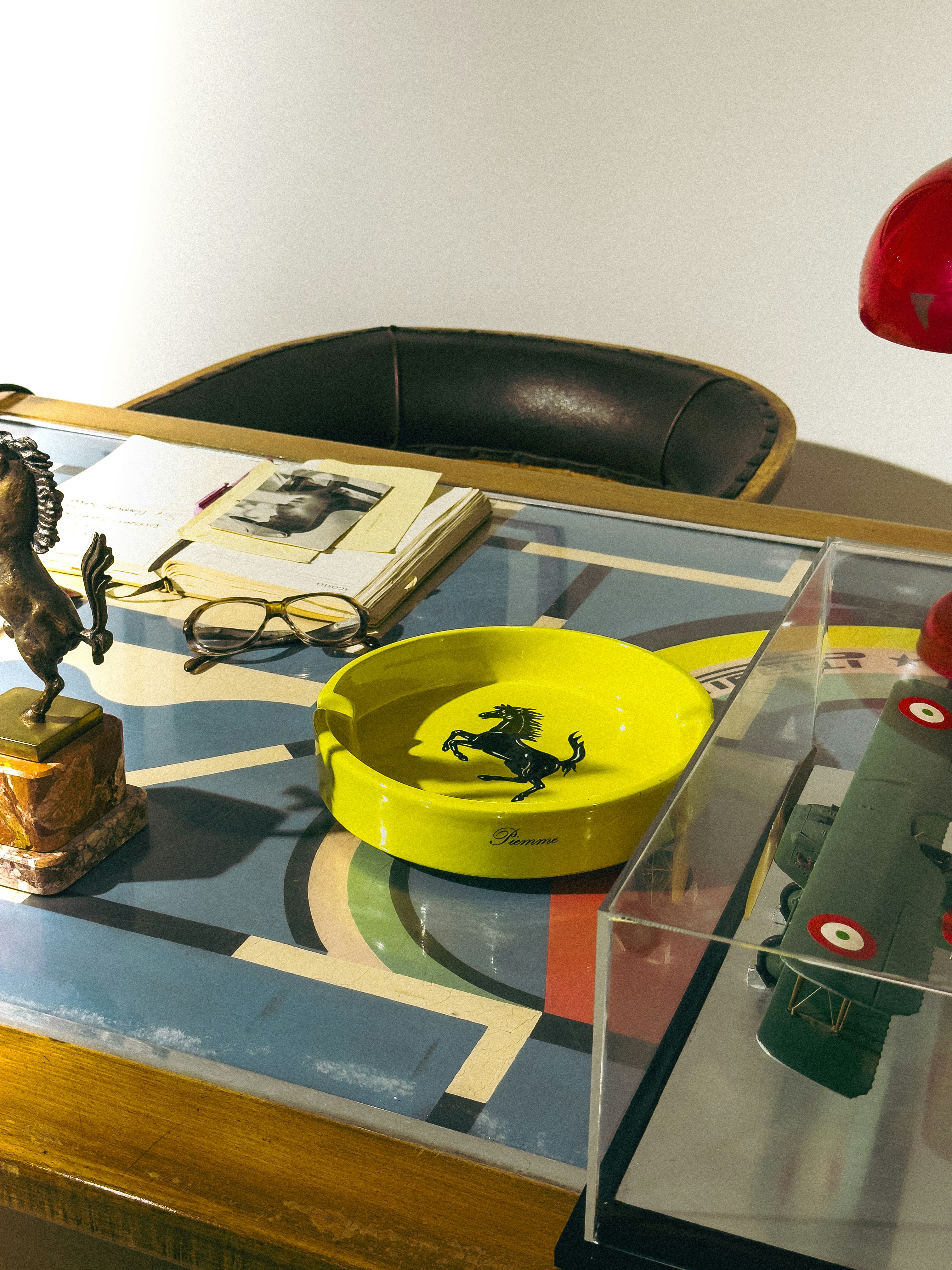 Ferrari Horse Logo in a Yellow Ashtray: A Glass-Desk Still Life with a Mini Plane and Vintage Accents