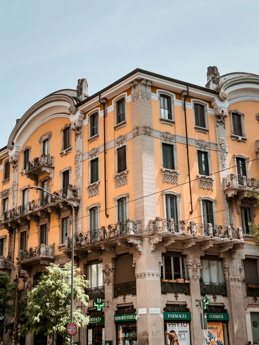 Sunlit ochre Art Nouveau corner building with ornate stone balconies and a street-level pharmacy.