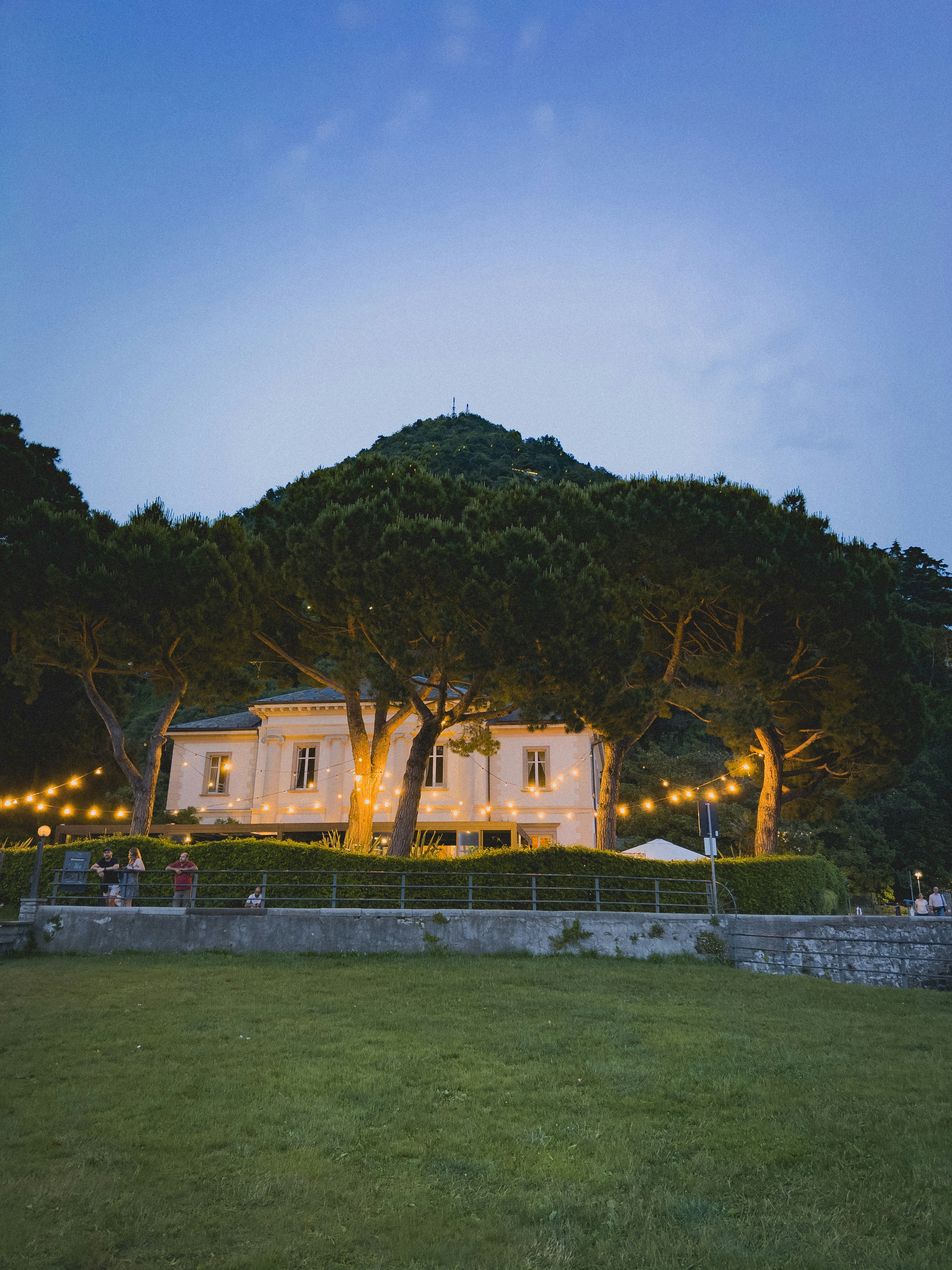 Charming villa illuminated by string lights at dusk near Lake Como, Italy. Surrounded by mountains and lush greenery, this serene evening scene captures the romantic essence of Italian lakeside living.