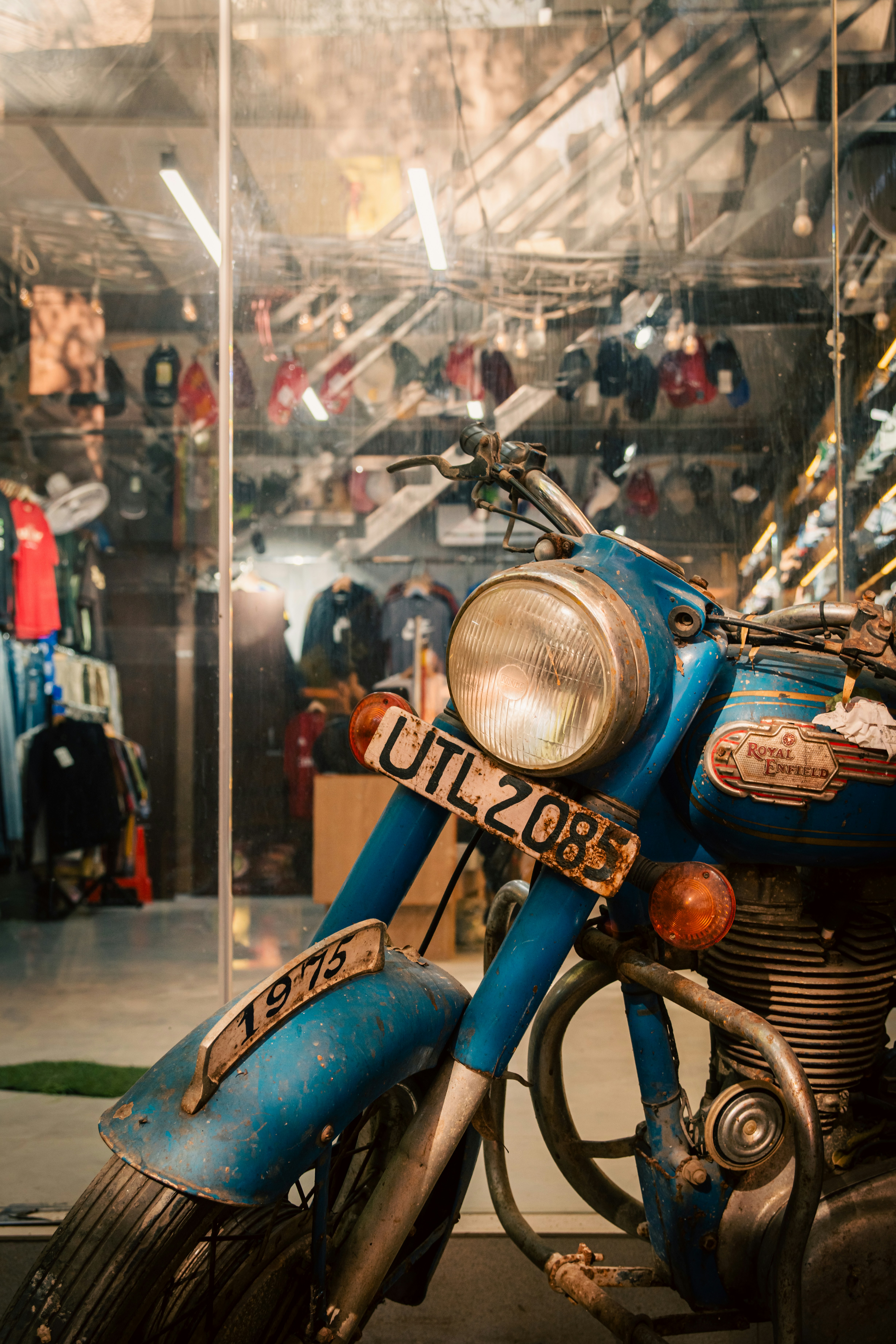 Timeworn Blue Motorbike Behind a Glass Storefront—Jackets and Neon Echo