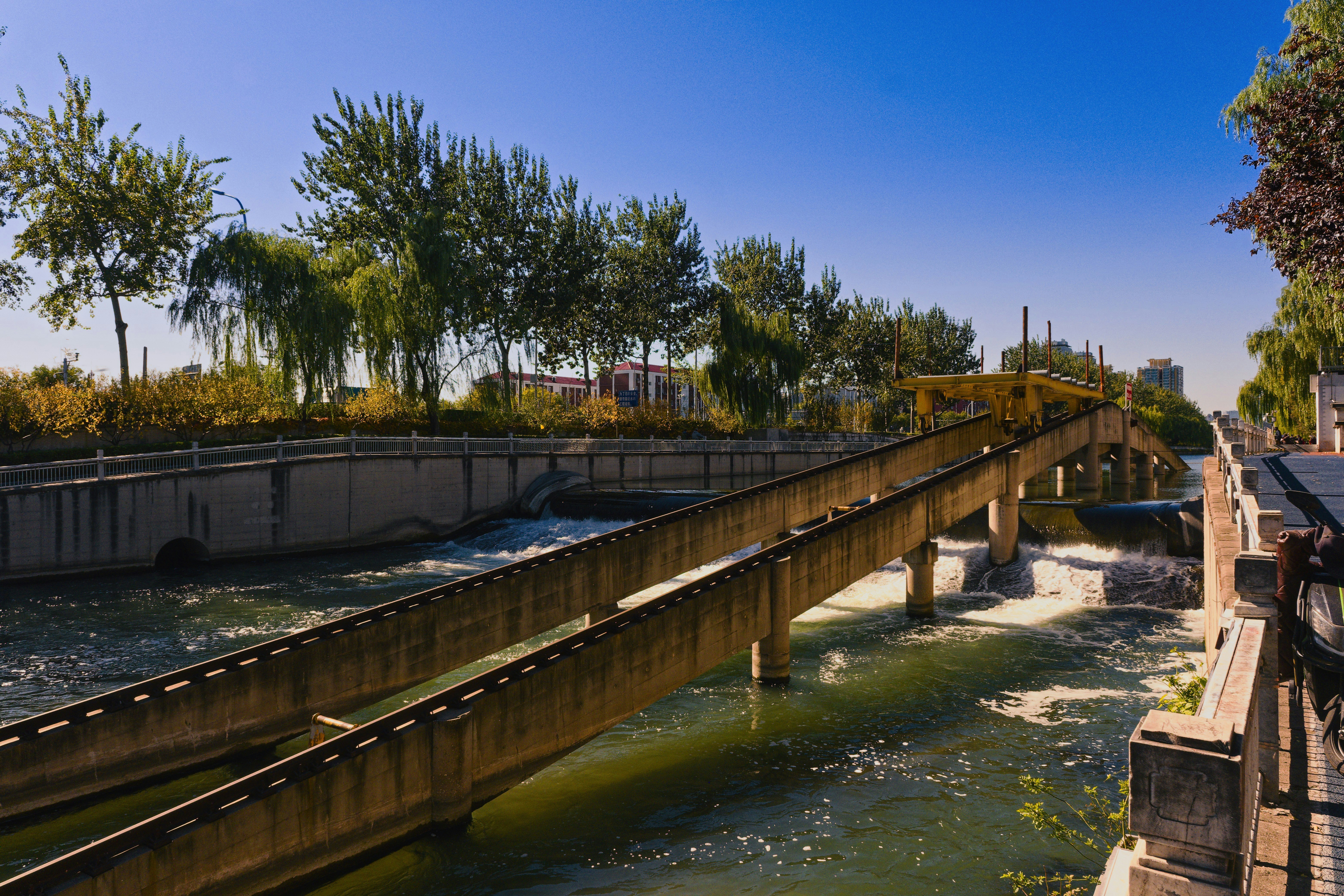 Bright sunny day at the urban waterway with scenic bridge and lush greenery in the background - A vibrant river scene shows a sunny day with clear blue skies. A sturdy bridge crosses the water, surrounded by leafy trees and urban structures, creating a peaceful atmosphere.