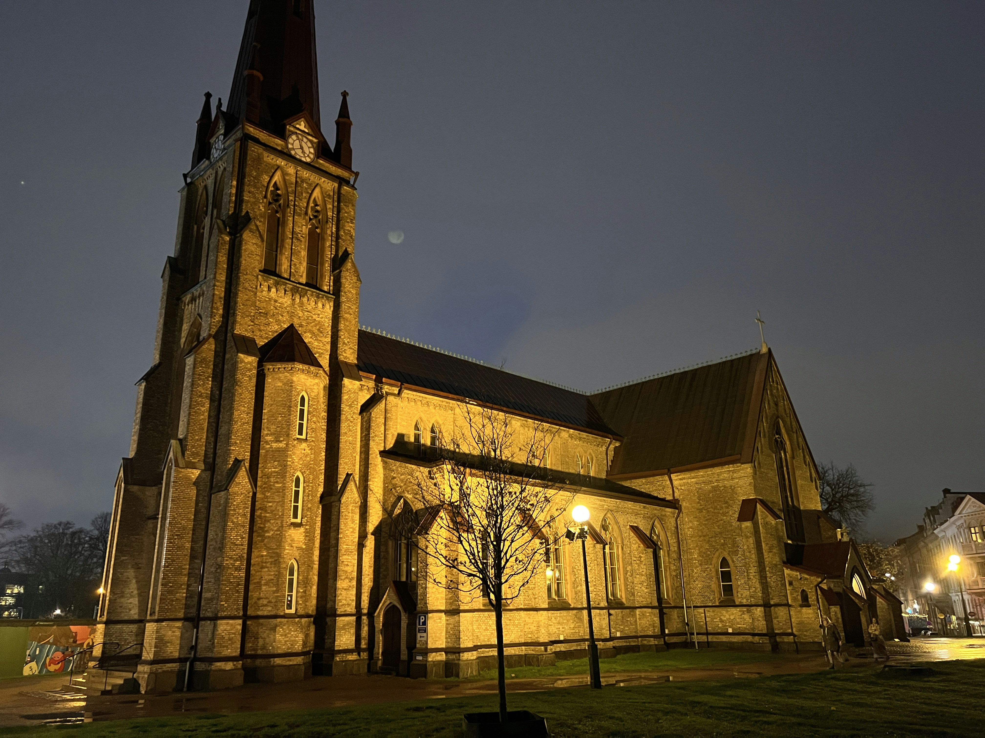 The majestic clock tower of a historic church stands sentinel at dusk, its façade aglow with warm lights amidst the tranquil Swedish night.