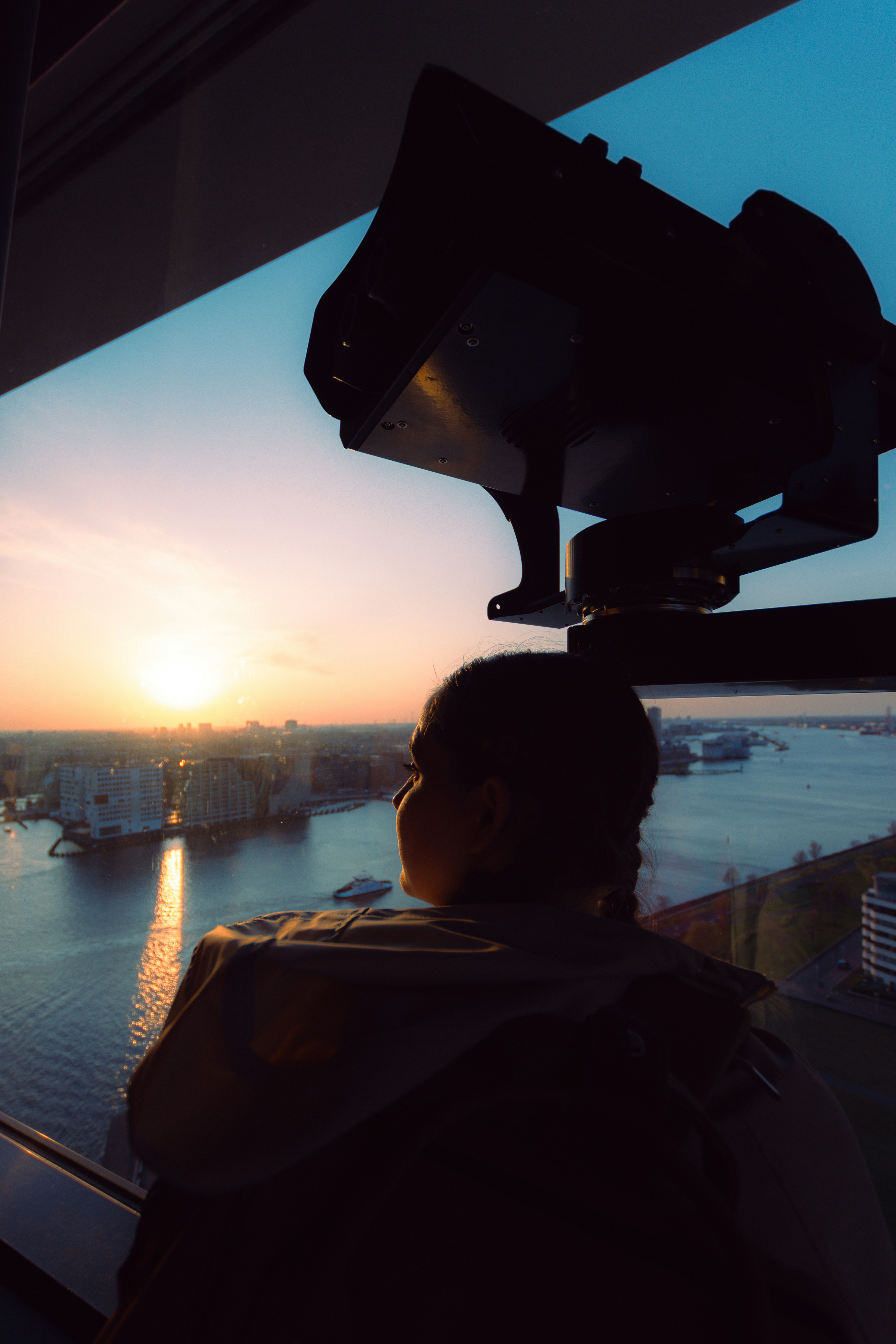 Le soleil se couche sur la ville et ma copine reste là, silencieuse, fascinée par la vue. La lumière dorée se reflète sur les vitres et l’eau, créant ce moment suspendu où tout paraît calme et infini.
