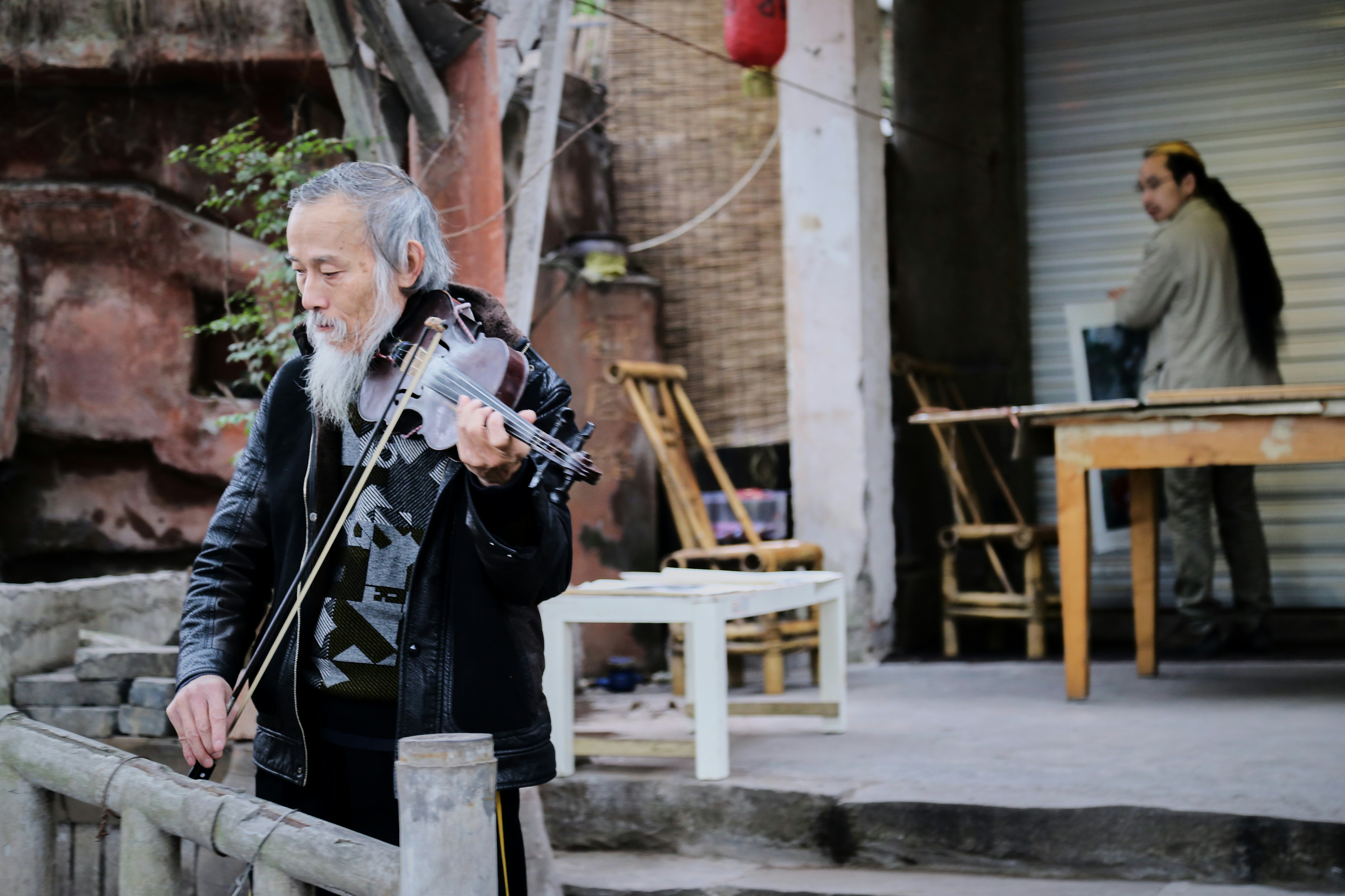 An elderly man plays the violin outdoors in a rustic courtyard, while another person works at a wooden table in the background. The scene feels intimate and artistic, capturing a quiet moment of music and craftsmanship.
