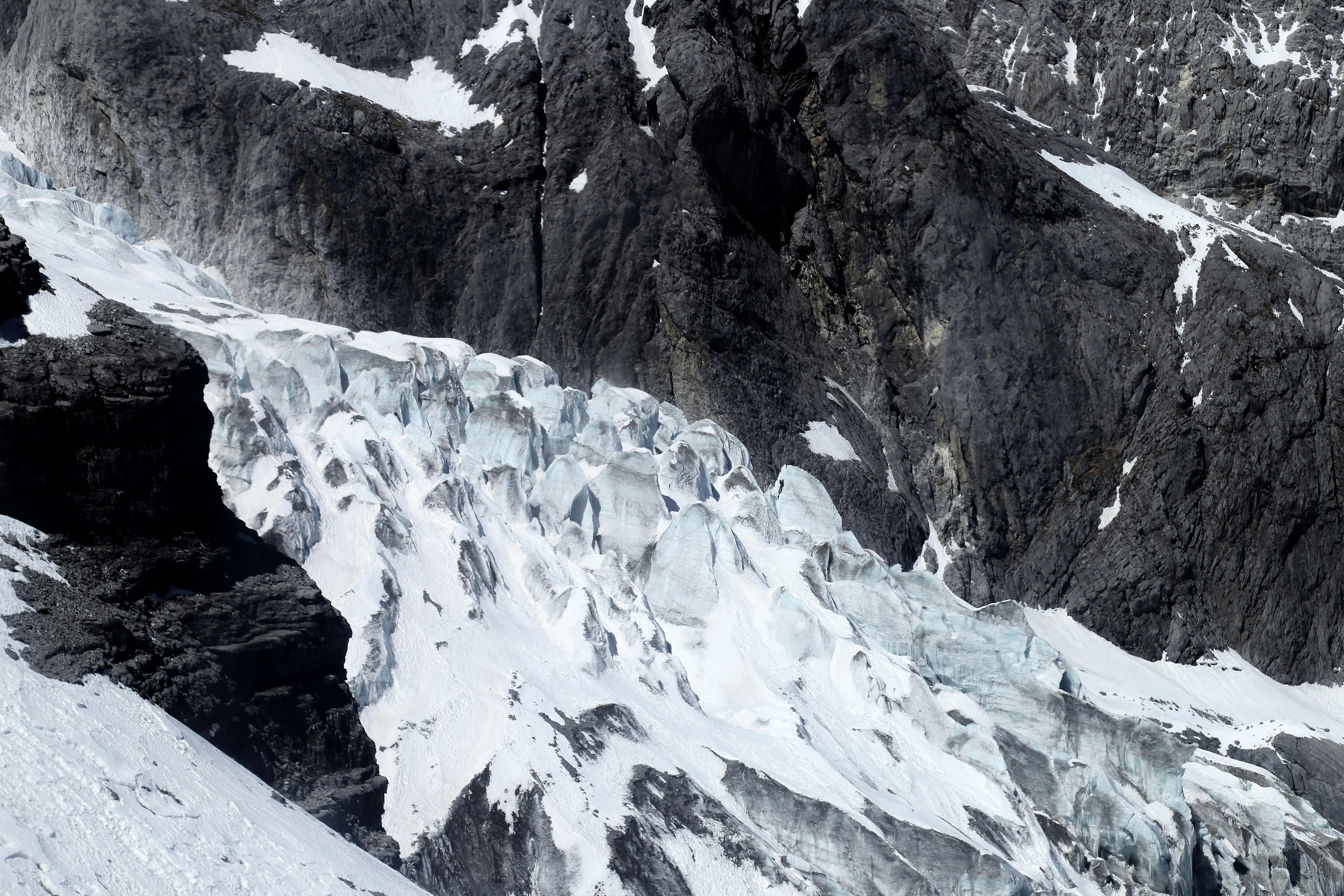 A dramatic view of a glacier flowing down steep dark rock cliffs, with layers of ice and snow forming sharp ridges and crevices. The contrast between the icy blue glacier and rugged mountains highlights the raw power of alpine nature.