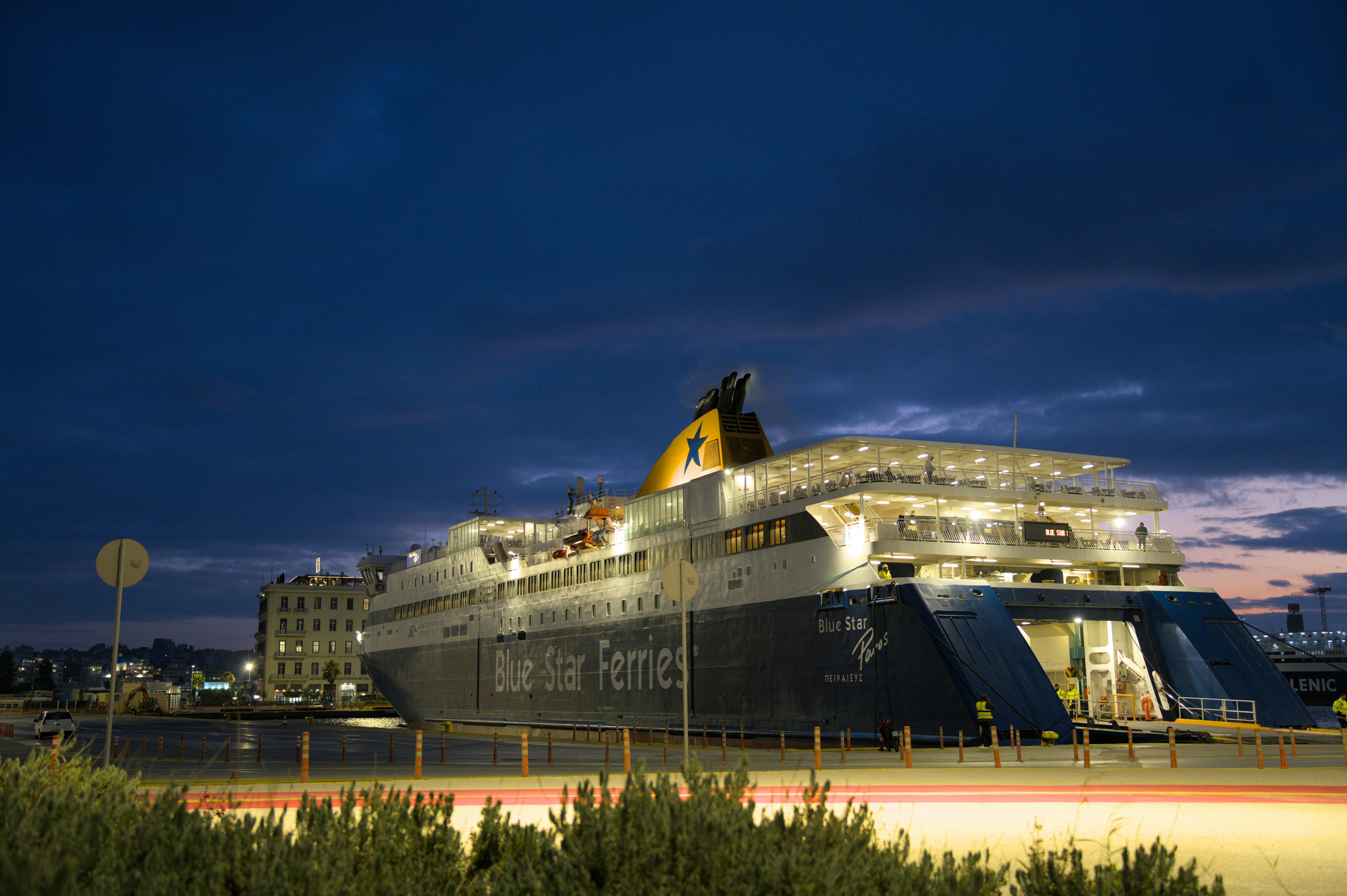 Nighttime photograph of Blue Star Ferries docked at the quay, with warm harbor lights outlining the vessel. Long exposure reveals light trails from traffic in the foreground.