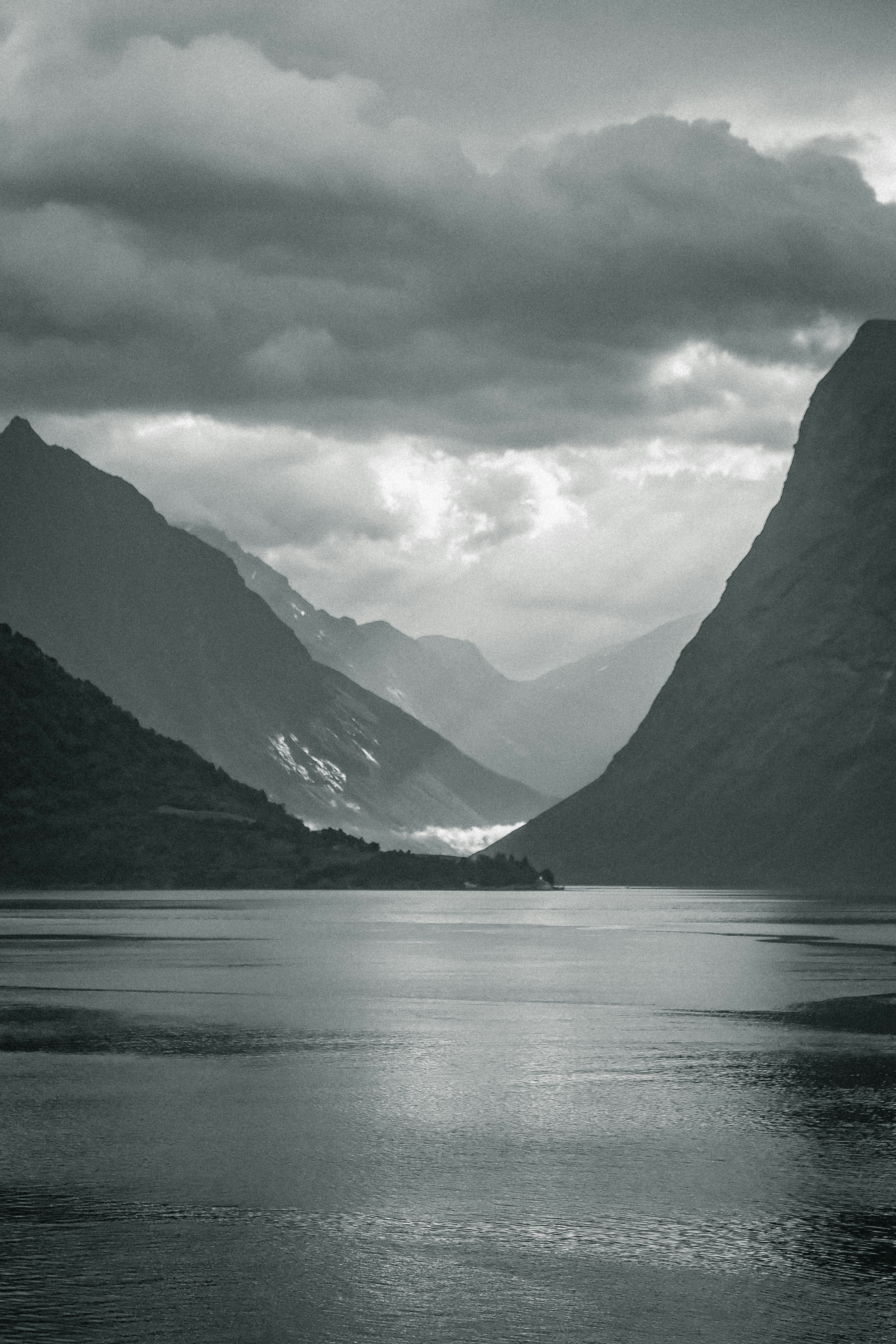 A timeless black and white photograph capturing the dramatic serenity of a Nordic fjord. Towering mountains flank the calm, reflective waterway under a moody, overcast sky.