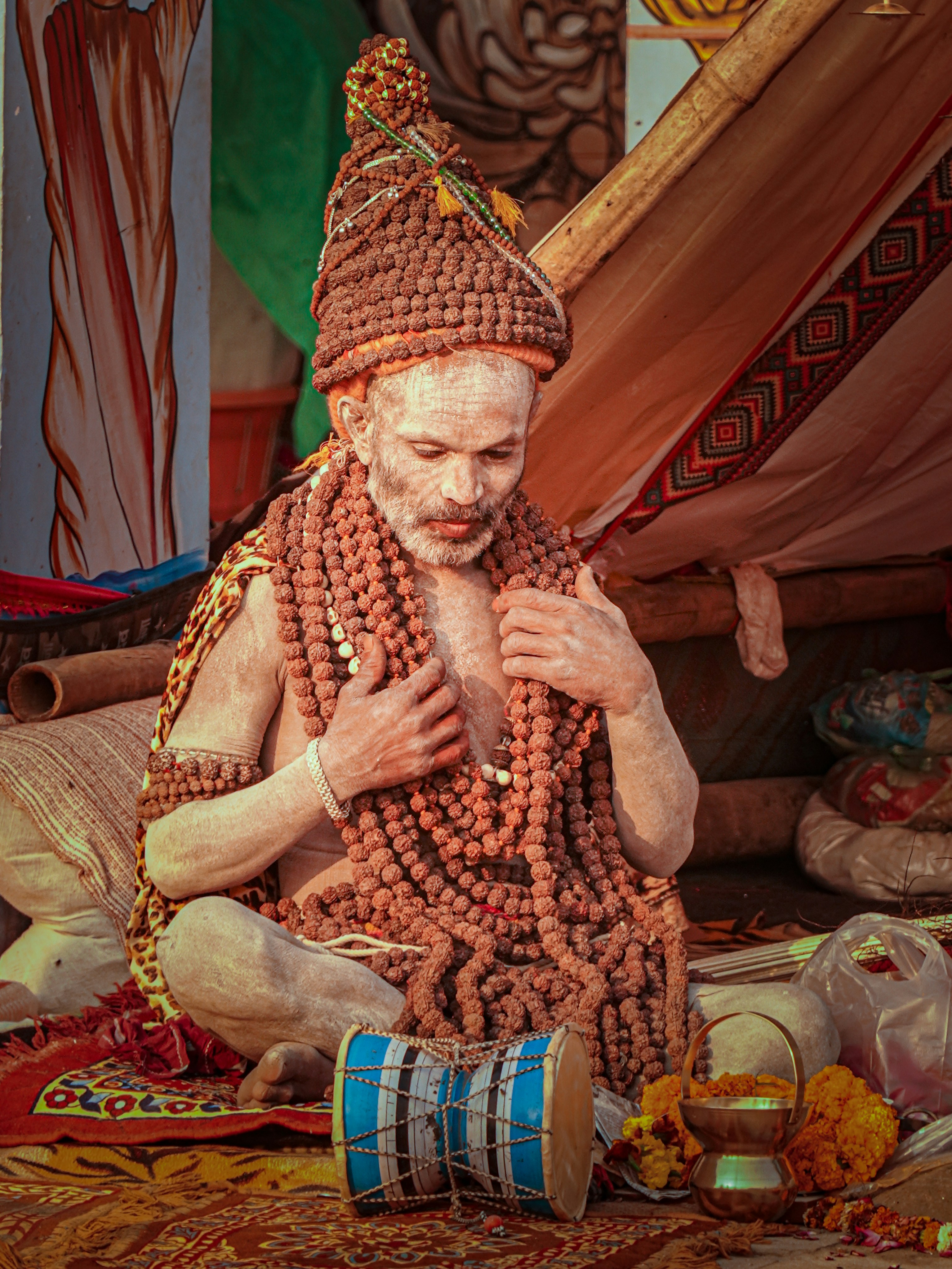 This is a powerful, intimate portrait of an Indian Sadhu (Hindu holy man), fully immersed in his spiritual practice. The man's body is entirely covered in Bhasma (sacred ash), symbolizing renunciation and his identity as a follower of Lord Shiva. His most striking adornment is the overwhelming collection of Rudraksha beads, which he wears as a tall, conical cap and cascading necklaces that cover his chest. These beads, said to be the tears of Lord Shiva, are sacred tools that enhance meditation, offer protection, and signify his deep connection to the divine.