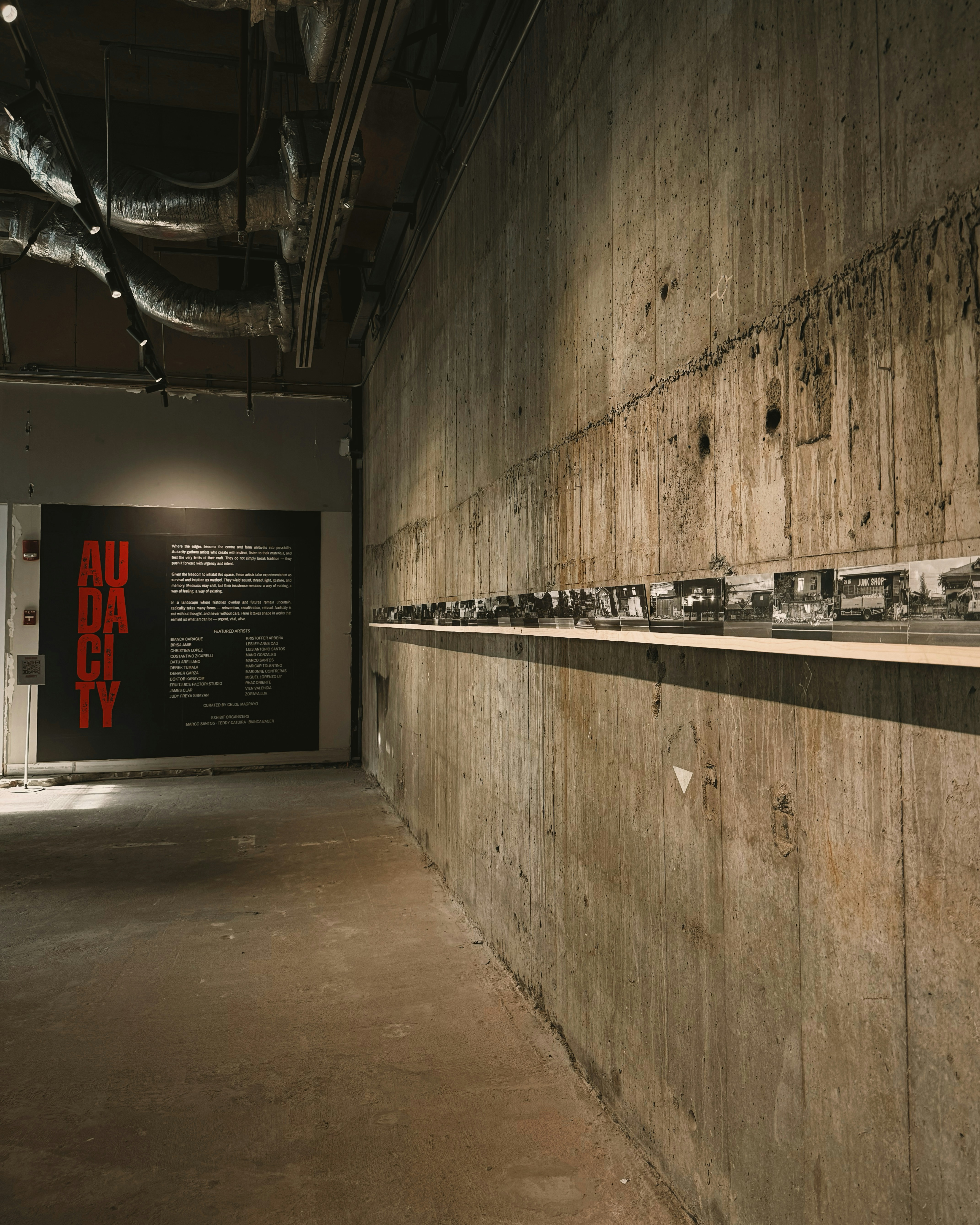 Dim industrial corridor with a long row of framed photos along the right wall and a bold red 'AUDACITY' sign on a black wall to the left.