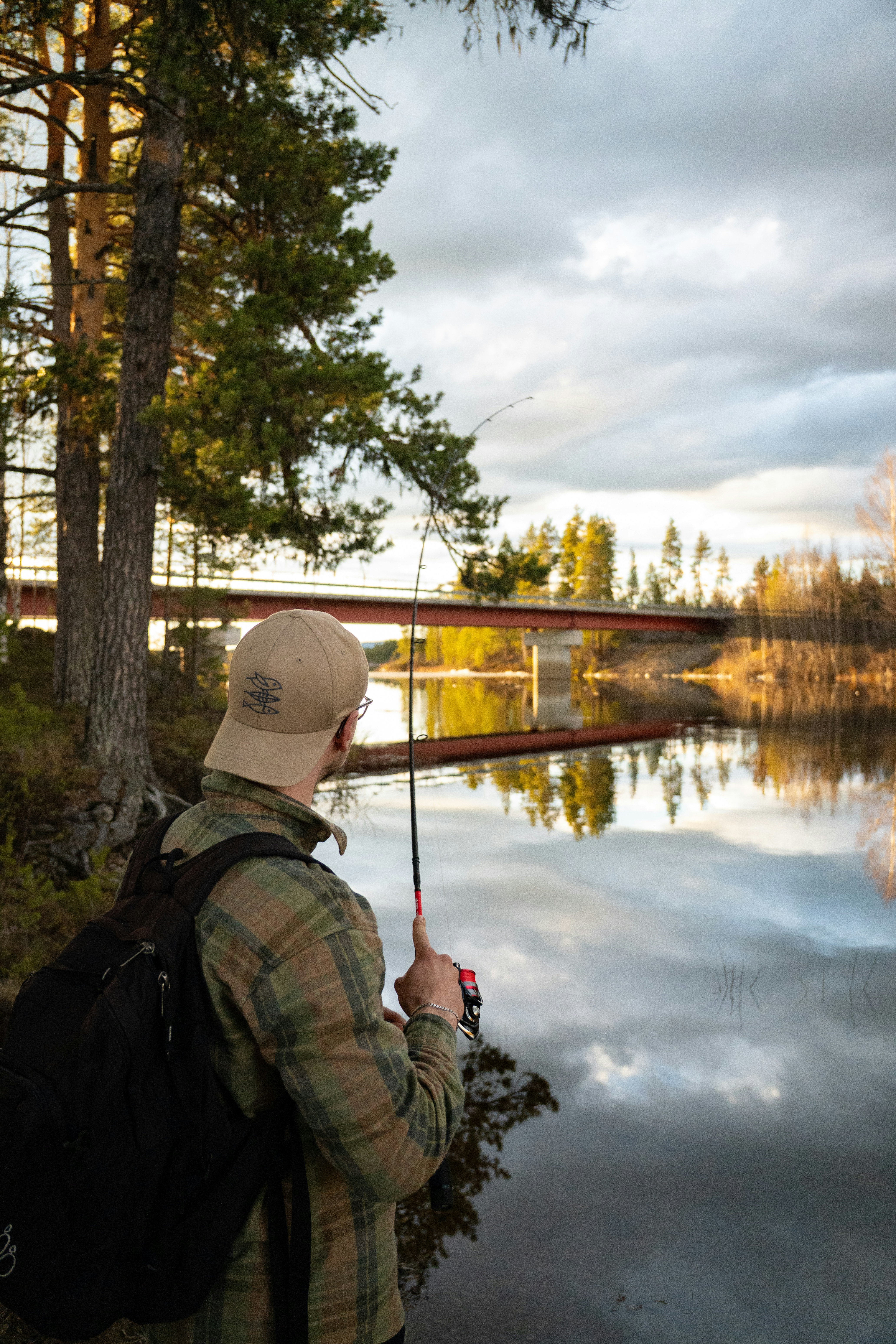 Casting Toward Reflection: A Lone Angler by a Tranquil Lake Beneath a Red Bridge