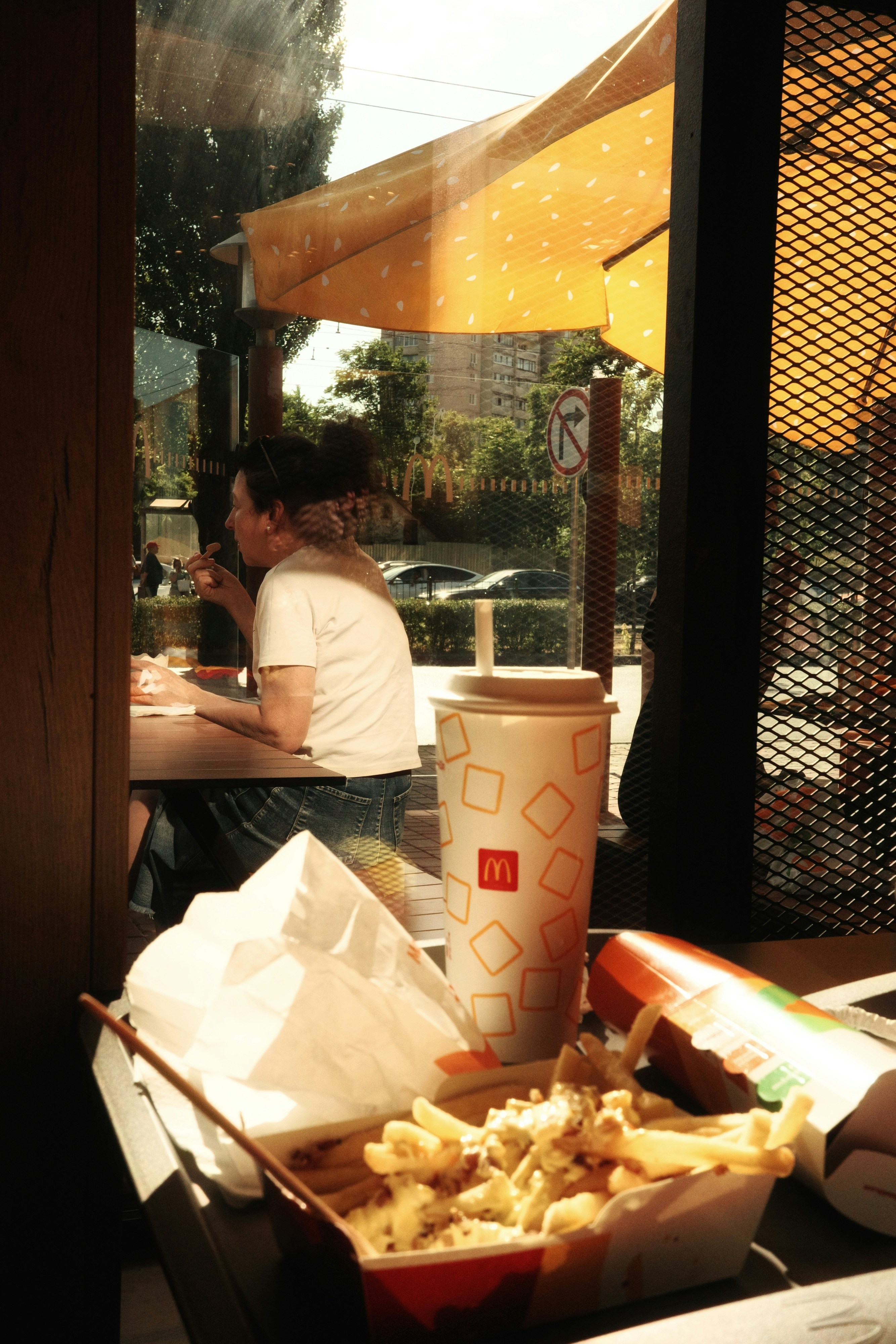 A warm, sunlit indoor shot captures a fast-food meal from the perspective of the table. In the foreground, a tray holds an open carton of French fries topped with cheese or sauce, a drink cup with the McDonald's logo, and wrappers. The light streams in from a large window on the left, where a woman is seated at a table, eating. Outside, a bright yellow awning patterned with white dots creates a strong visual element, and the sunny view beyond shows trees, parked cars, and a red traffic sign with a diagonal slash. The image juxtaposes the quick, indoor meal with the bright, busy world outside.