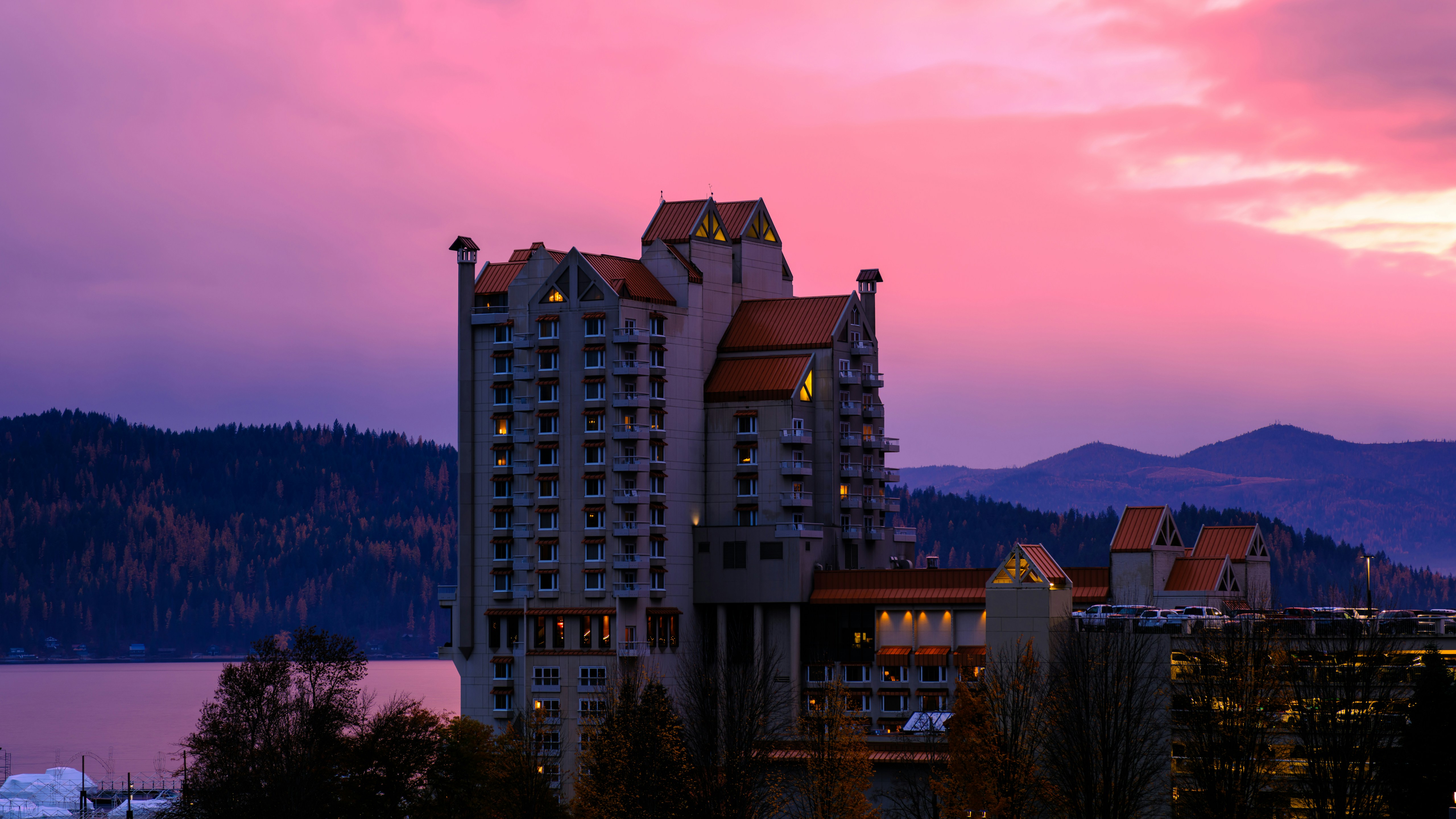 A serene sunset over Coeur d’Alene with warm pink and purple skies illuminating the lake and surrounding mountains. The Coeur d’Alene Resort rises against the calm evening backdrop, its windows glowing as dusk settles over northern Idaho.