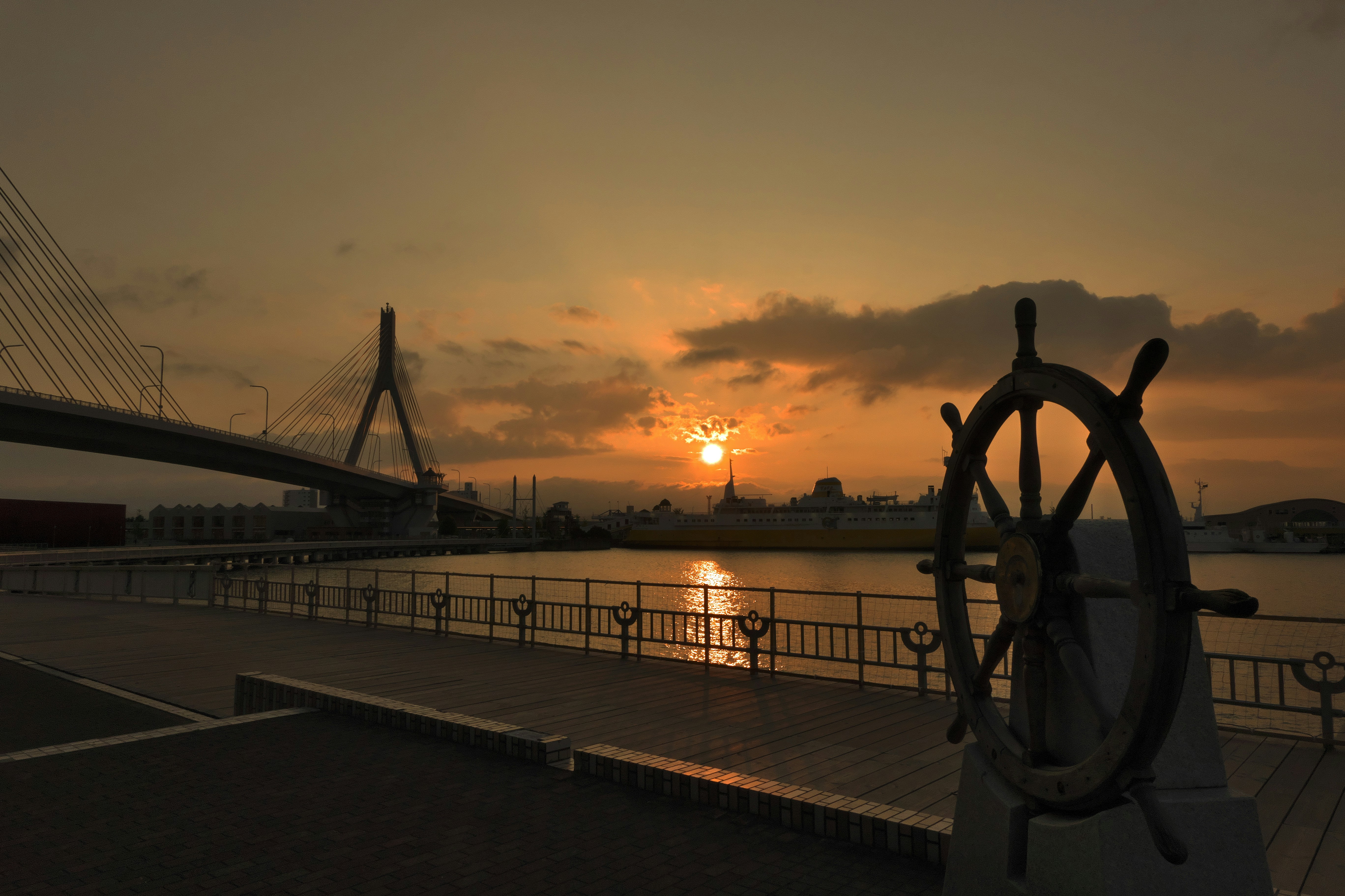 Dusk at the Harbor: A Ship's Wheel Framing Sunset Over a Quiet Riverfront