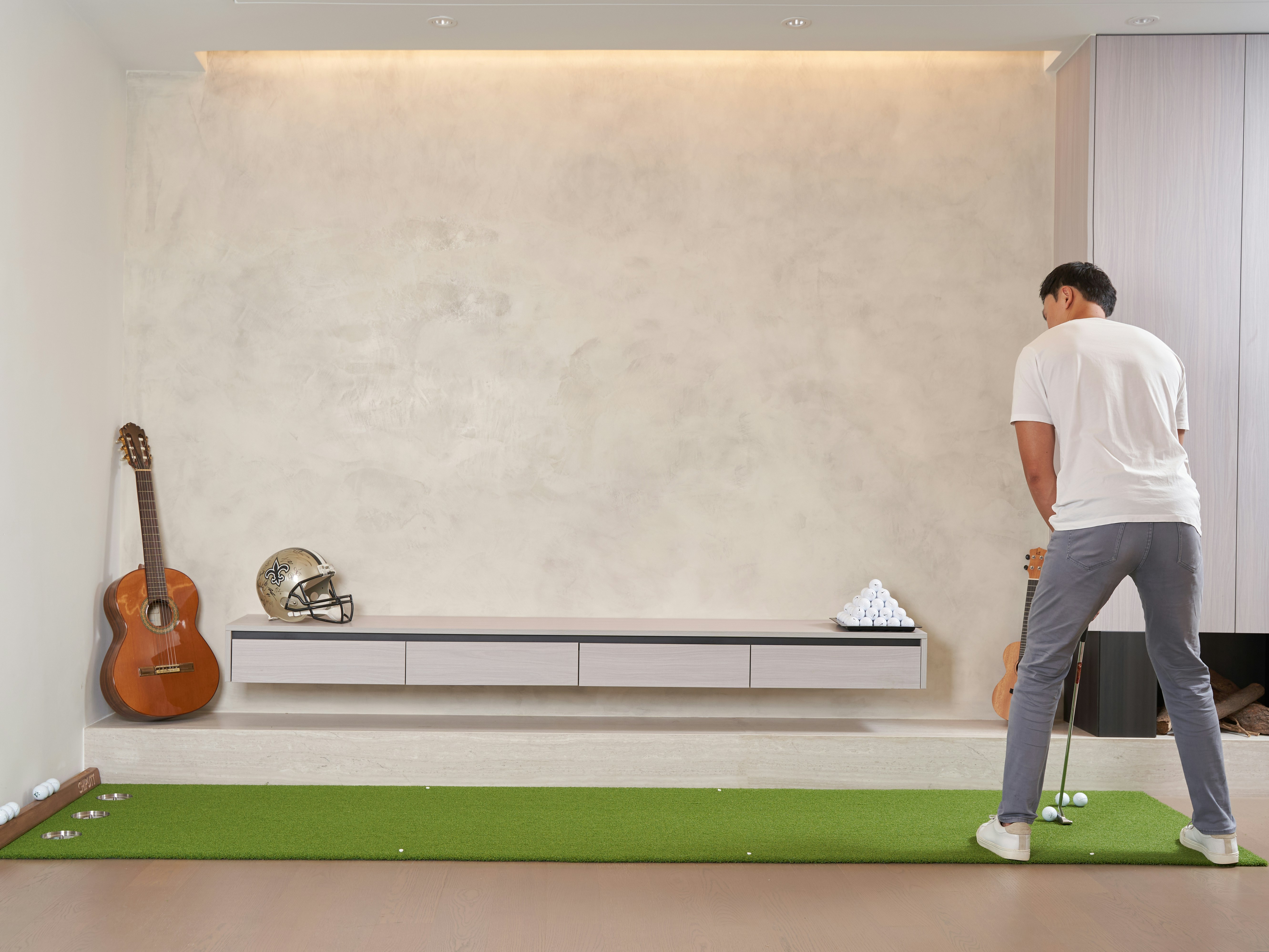 Image by Chiputt Golf. Learn more at TheChiputt.com. A golfer practices in a bright, minimalist living room, standing over a long strip of indoor turf laid across the floor. A guitar, a football helmet, and a stack of golf balls rest along the walls, adding personal touches to the quiet, relaxed space. Soft lighting and simple décor create a peaceful atmosphere that blends everyday home life with the steady rhythm of golf practice.