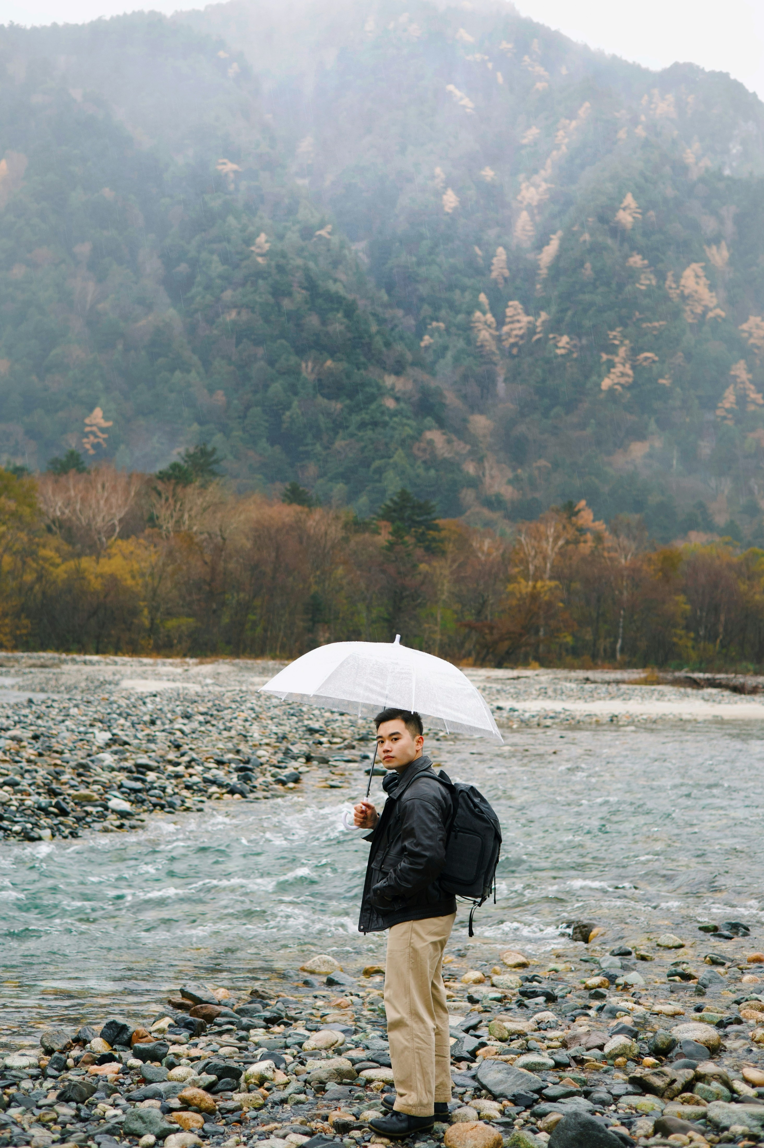 Umbrella at the Riverbank: A solitary figure framed by autumn forest and misty mountains