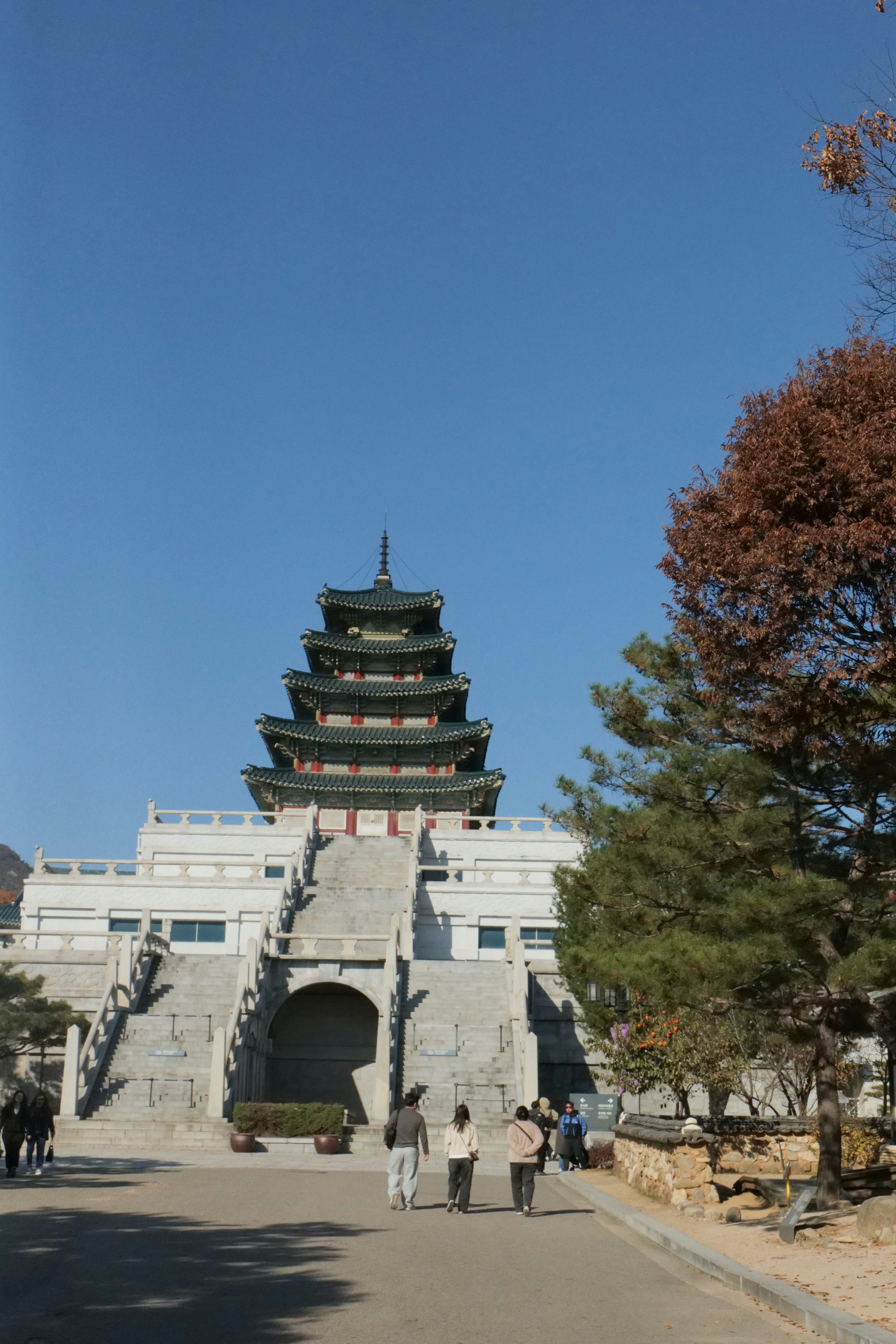 Autumn-colored pagoda rises above a sweeping stone staircase as visitors approach beneath a clear blue sky.