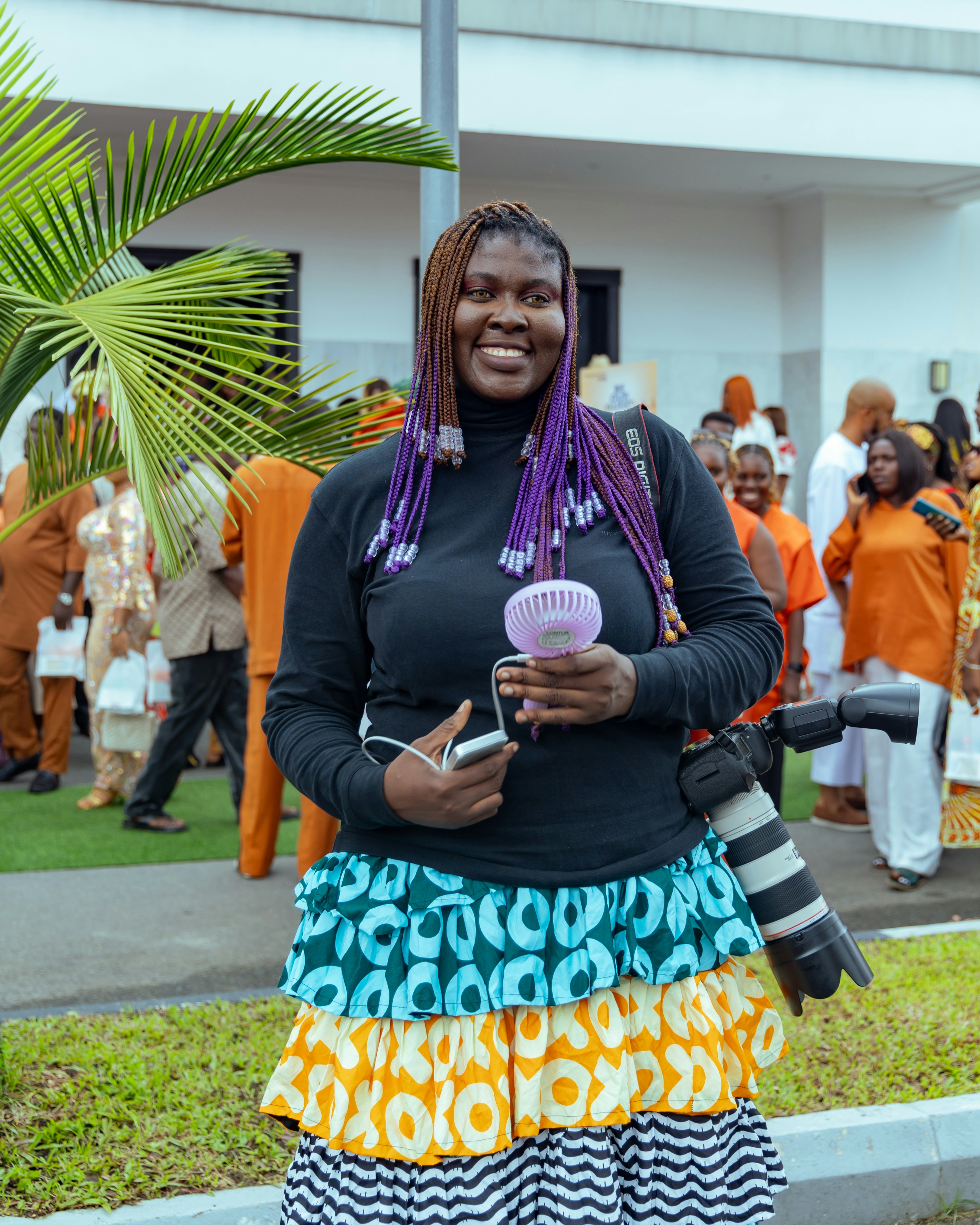 Candid portrait of a smiling woman in a black top and layered turquoise-yellow skirt at an outdoor event, holding a pink handheld fan and a telephoto lens.