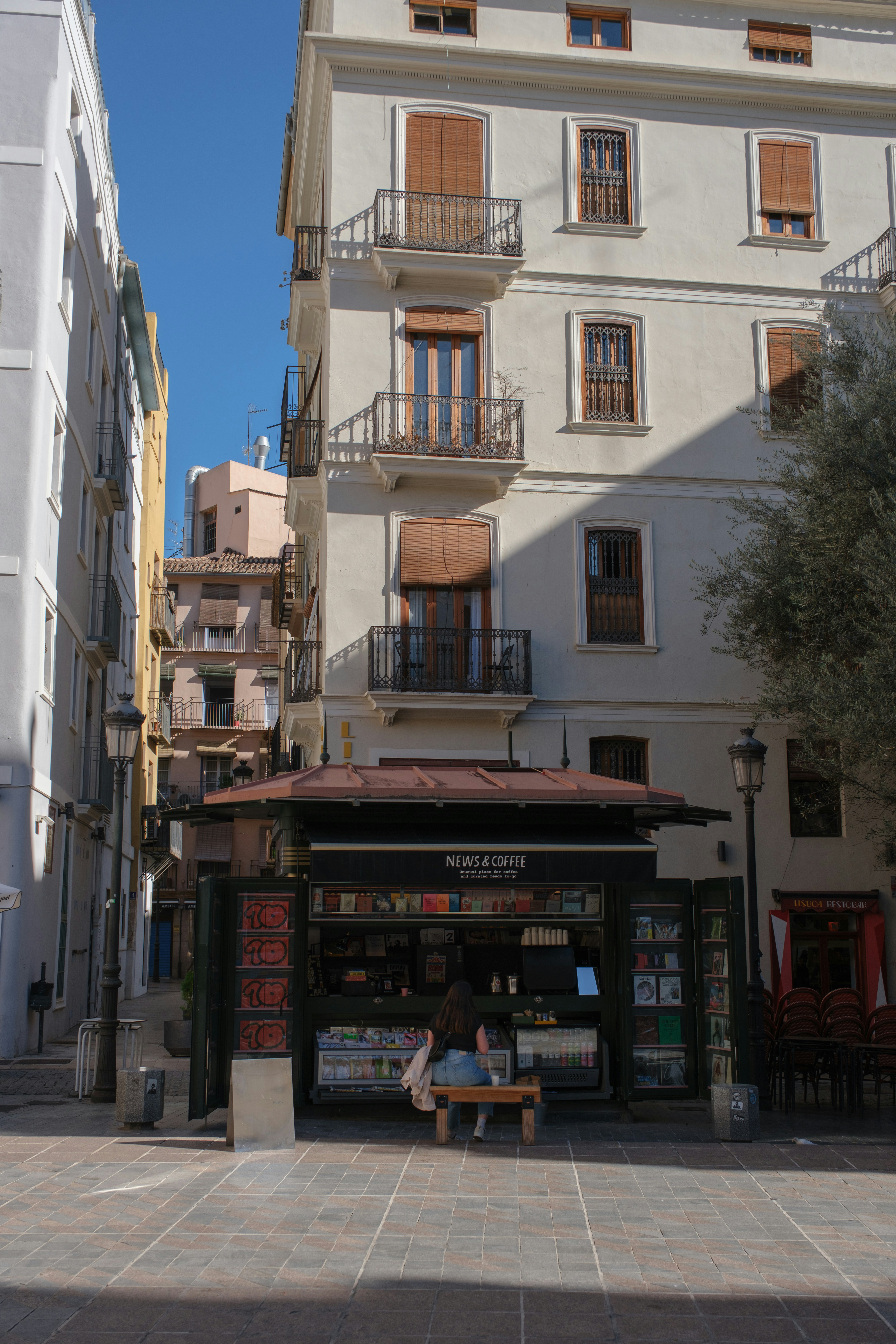 Sunlit News & Coffee Kiosk Beneath a Balcony-Lined Facade