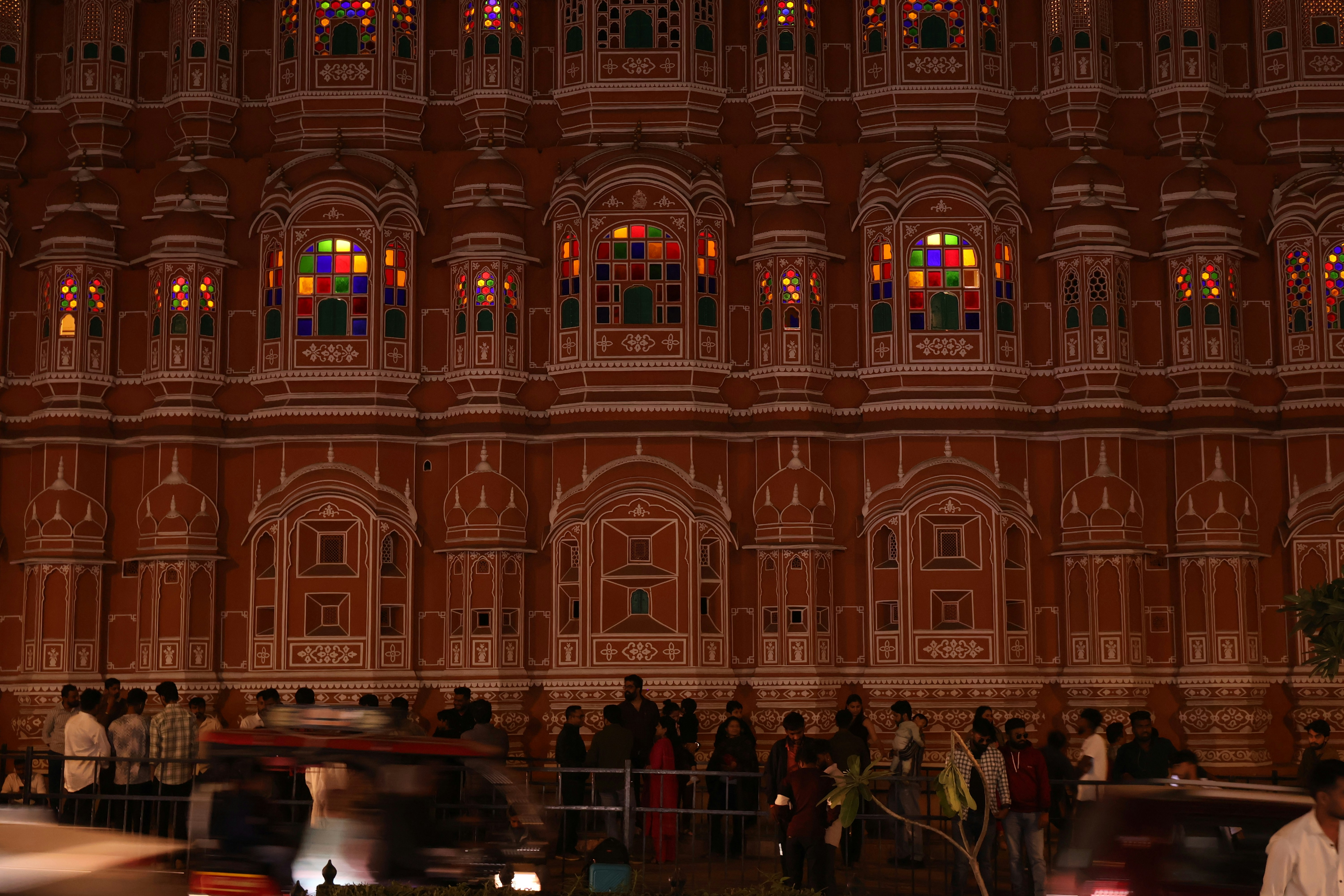The busy street in front of Hawa Mahal in Jaipur, India bustling with crowd taking pictures in front of the palace facade. The facade is dimly lit in night and has backlit colorful windows.
