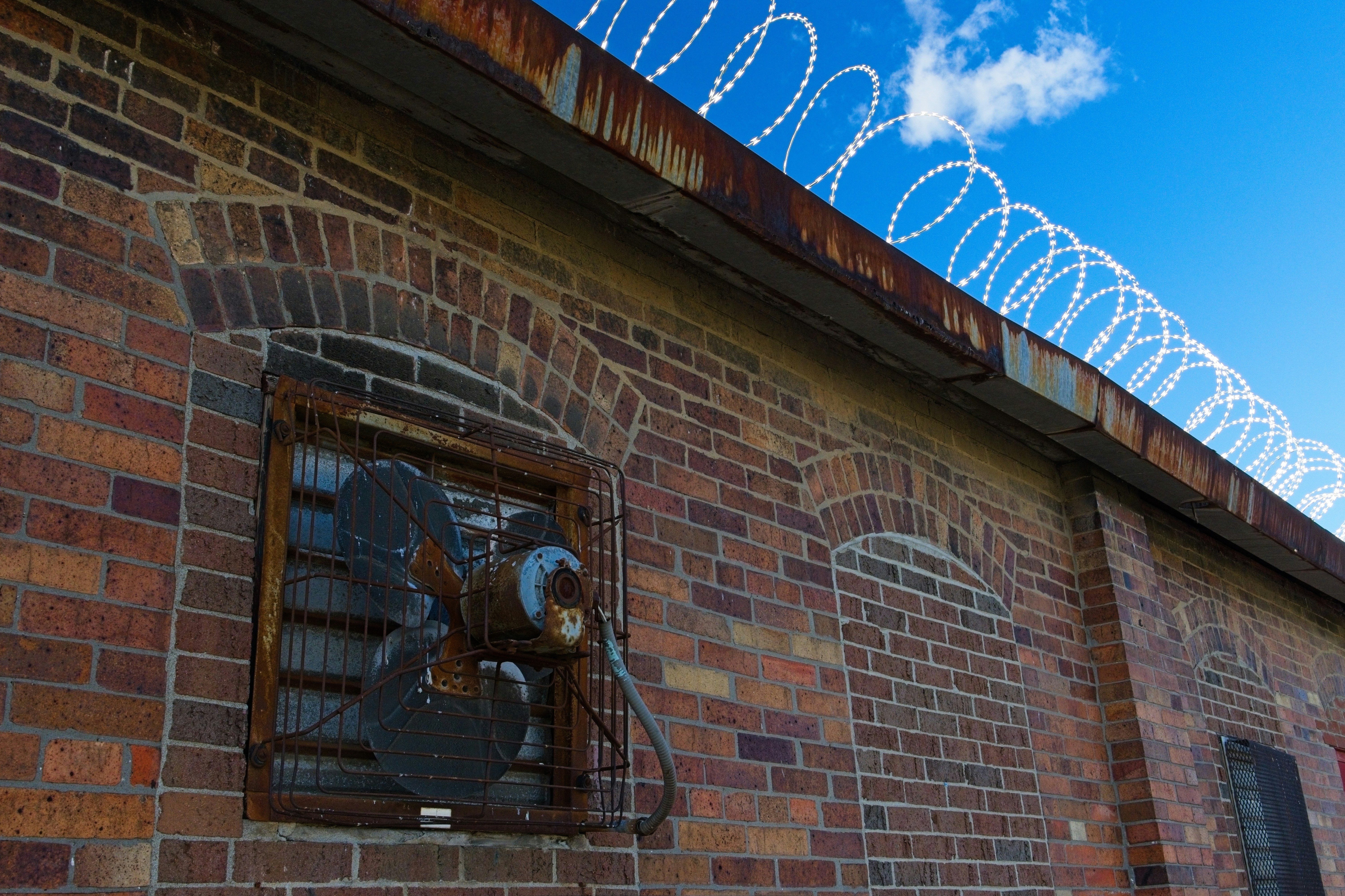 An old rusty exhaust fan hangs on a weathered brick wall with razor wire loops strung overhead.
