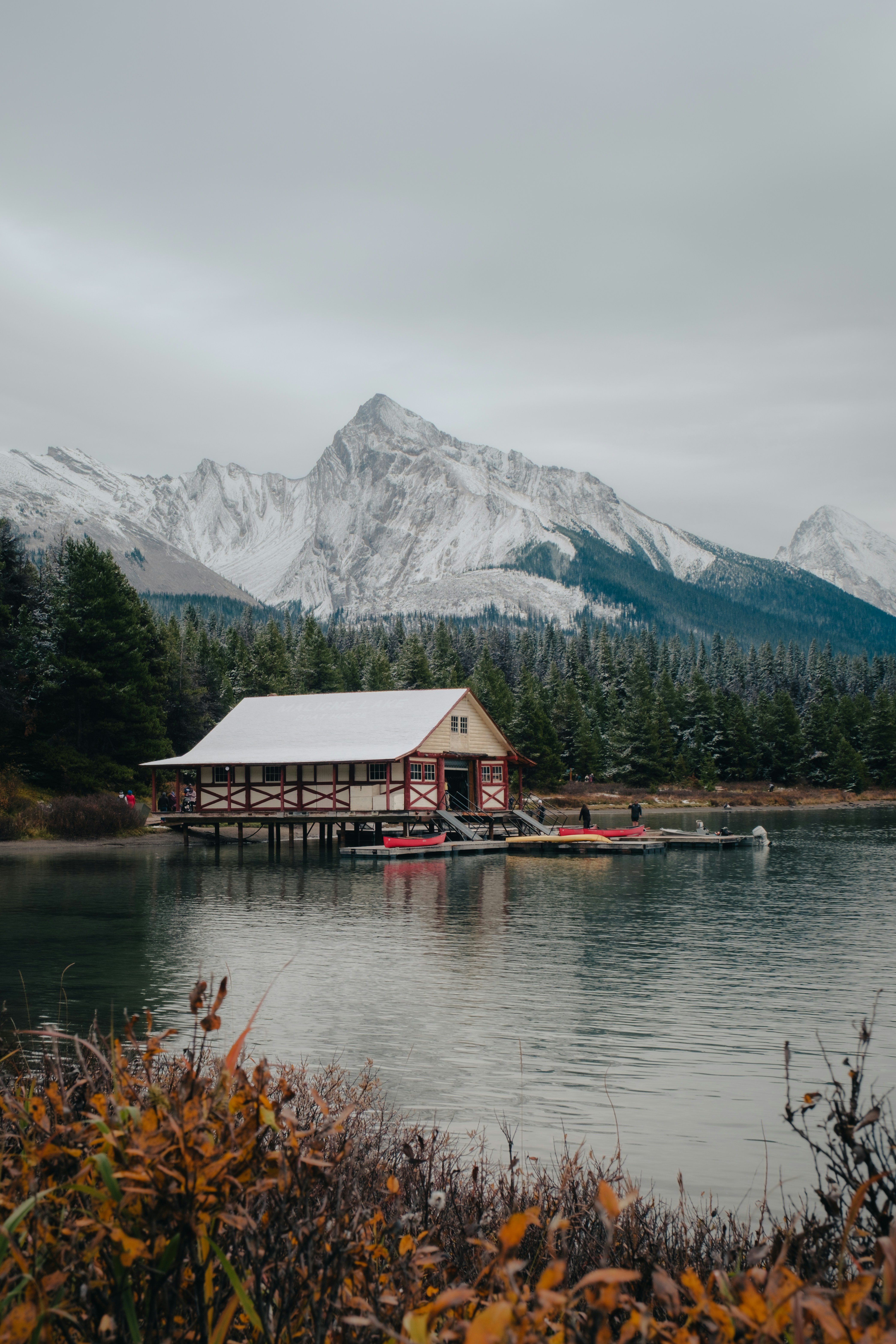 Alpine calm in the Canadian Rockies: a red-trim boathouse sits on a teal mountain lake beneath dark pines and fresh snow on the peaks. Soft overcast light and late-season foliage create a quiet, moody landscape in Alberta, Canada.