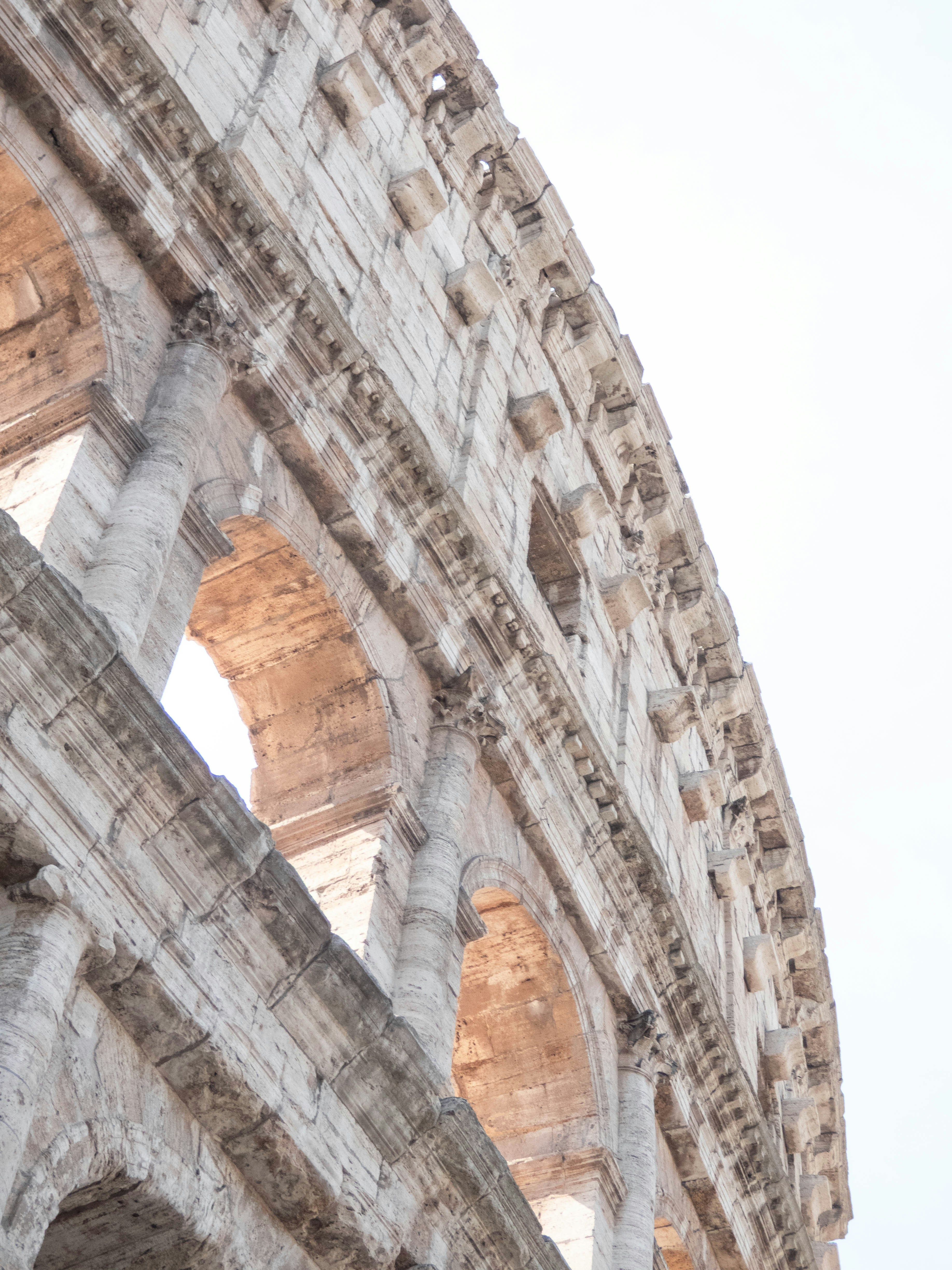 A close view of the Colosseum in Rome shows its stone arches and columns extending upward. The image captures multiple levels of the exterior structure with visible openings and architectural details.