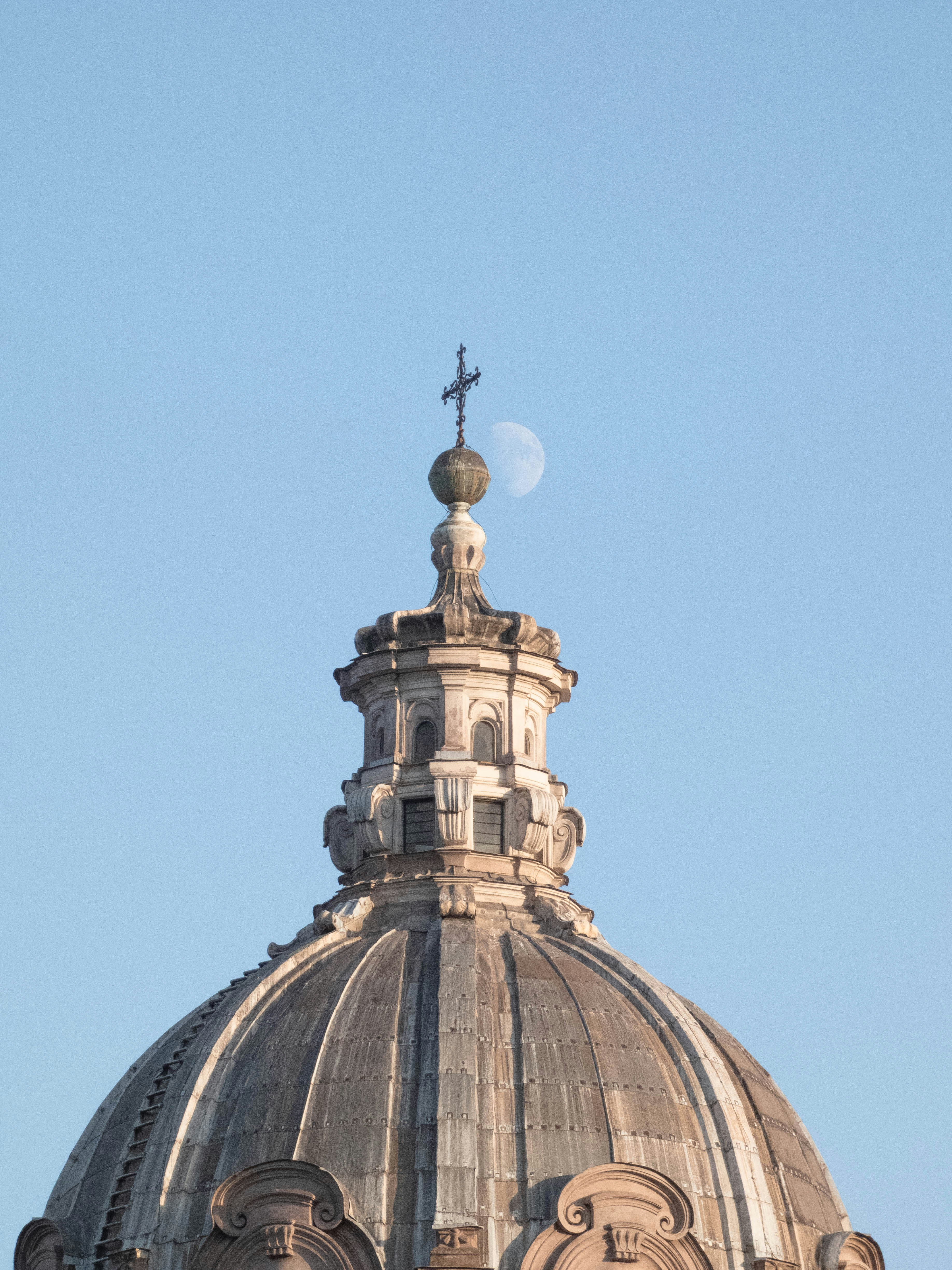 The upper section of a church dome topped with a cross is shown against a clear blue sky, with the moon visible nearby. The photo was taken outdoors in Rome, Italy.