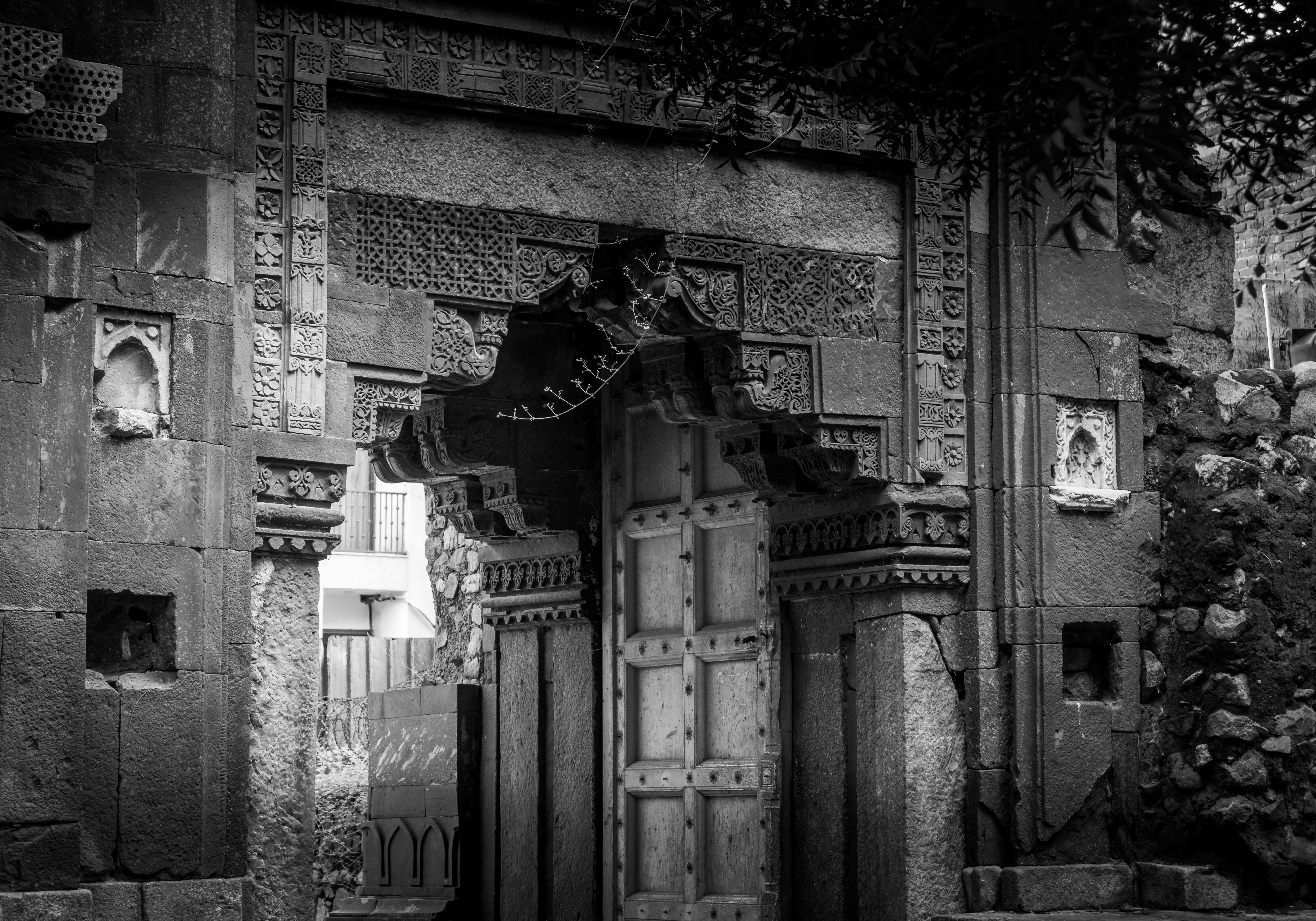 This photograph captures the richly carved entrance of Masjid Moth in South Delhi. The gateway is built from red sandstone and granite and features intricate Lodi period ornamentation. Decorative brackets, floral patterns, and recessed niches frame the tall wooden door at the centre. The weathering on the stone reveals centuries of exposure while the surrounding walls show a blend of rubble masonry and detailed craftsmanship.