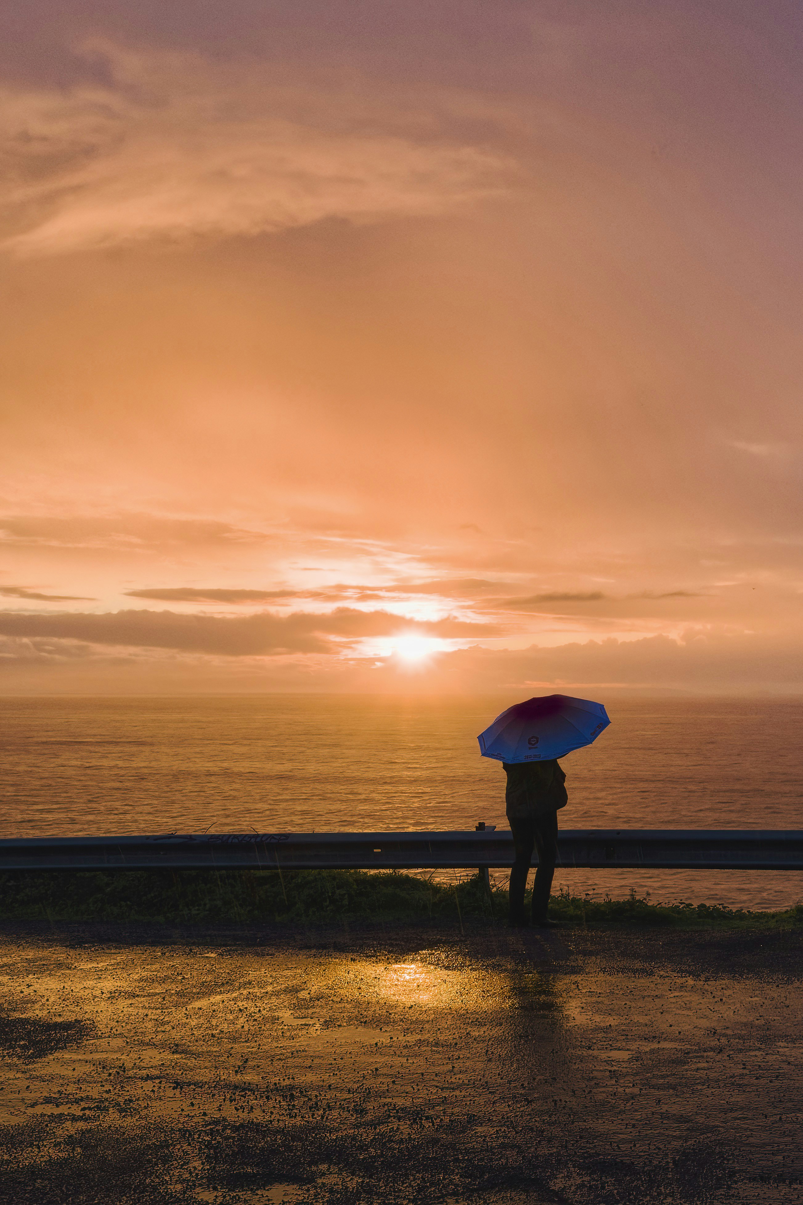 Person stands by the ocean under an umbrella watching a sunset with vibrant colors reflecting on the water - A person watches the sunset over the ocean while holding an umbrella. The sky is filled with warm colors, creating a beautiful and serene atmosphere.